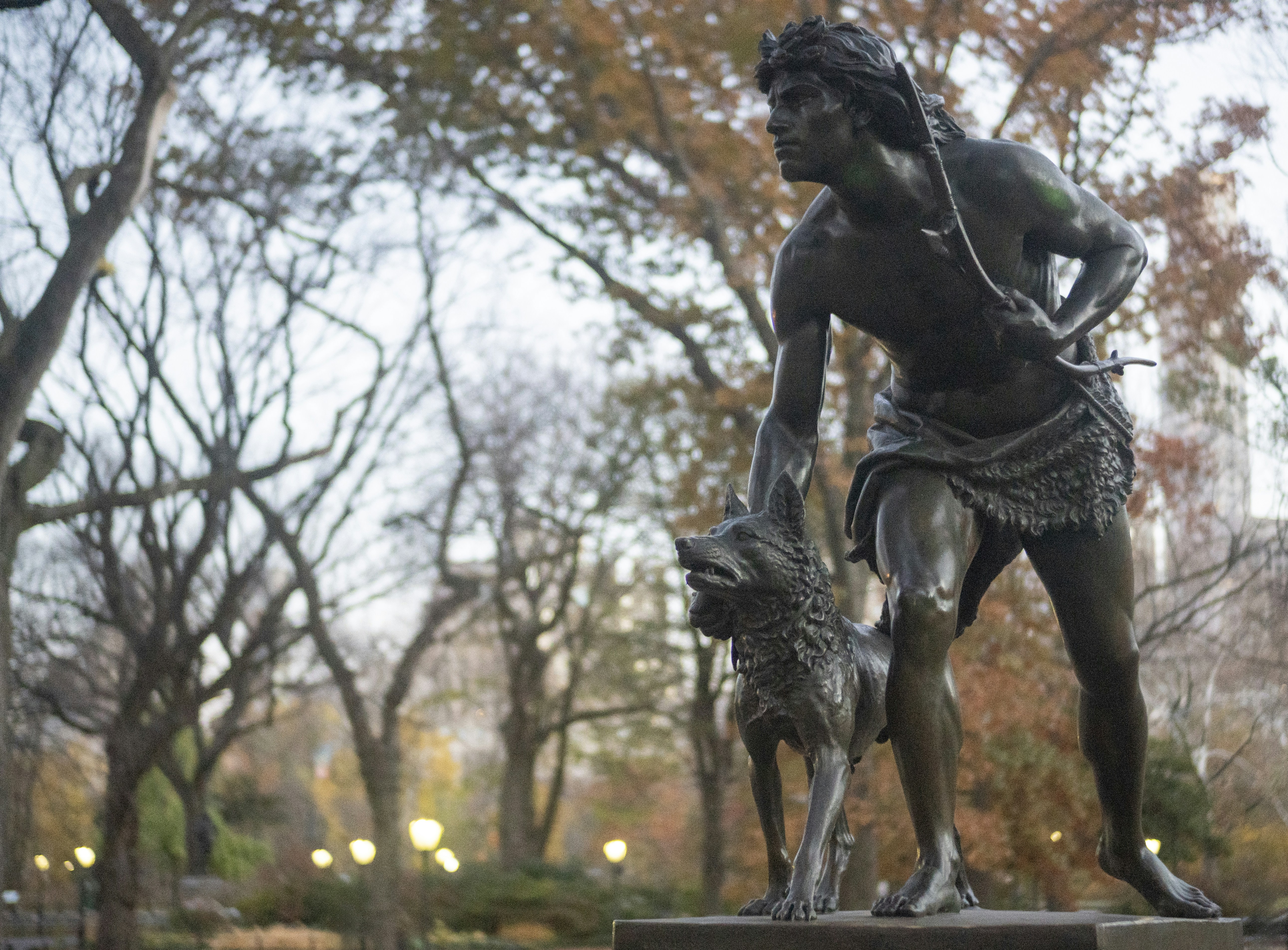 Bronze statue of a Native American figure with a wolf in a tree-lined park.