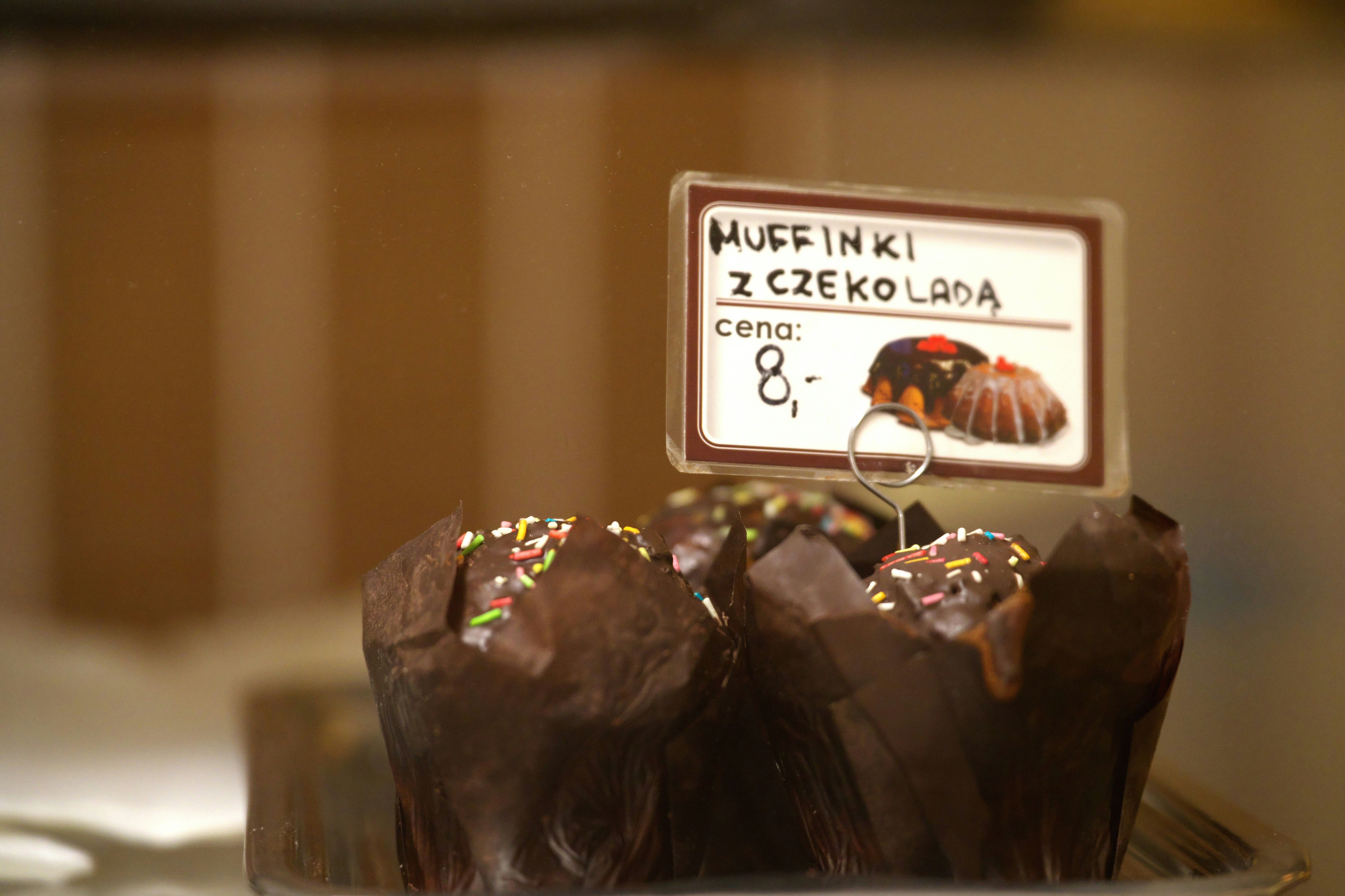 Chocolate muffins adorned with colorful sprinkles, displayed behind a glass case with a price tag. Perfect for dessert lovers.