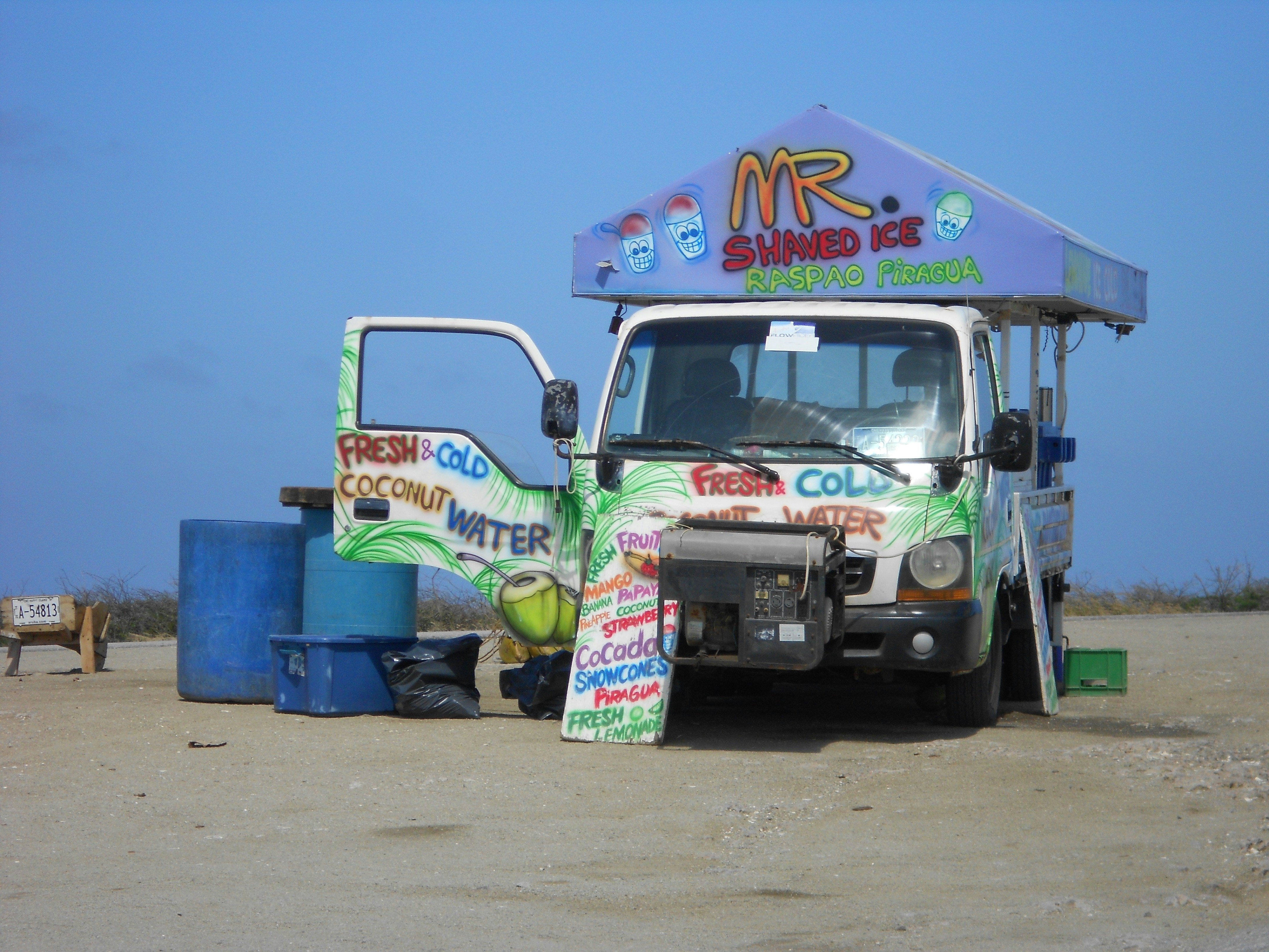 Colorful ice-cream vending truck parked on a sandy beach, its sides covered in hand-painted signage. The scene centers on the mobile cart against a clear sky.