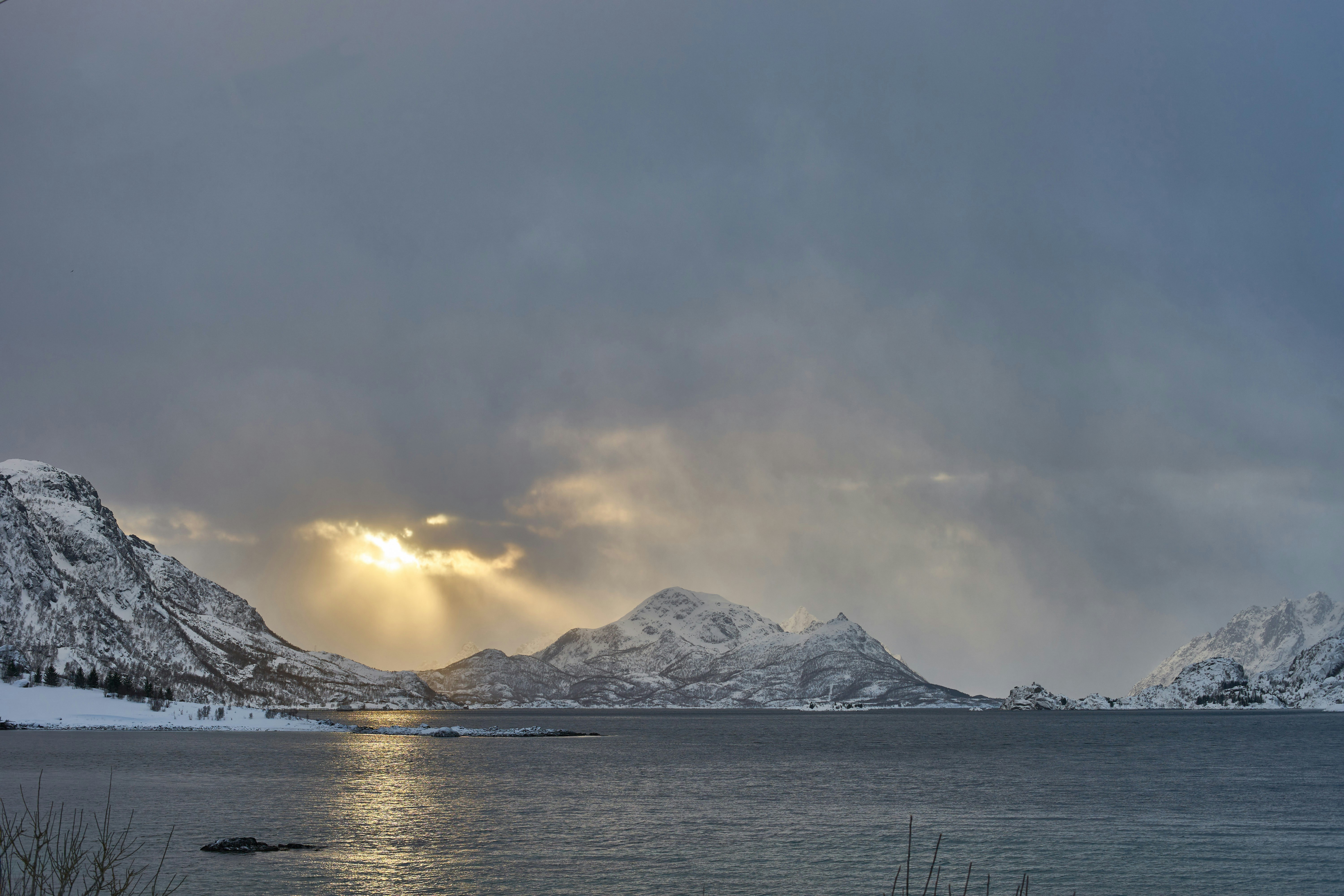 Sunlight breaks through clouds over snowy mountains and a calm sea.