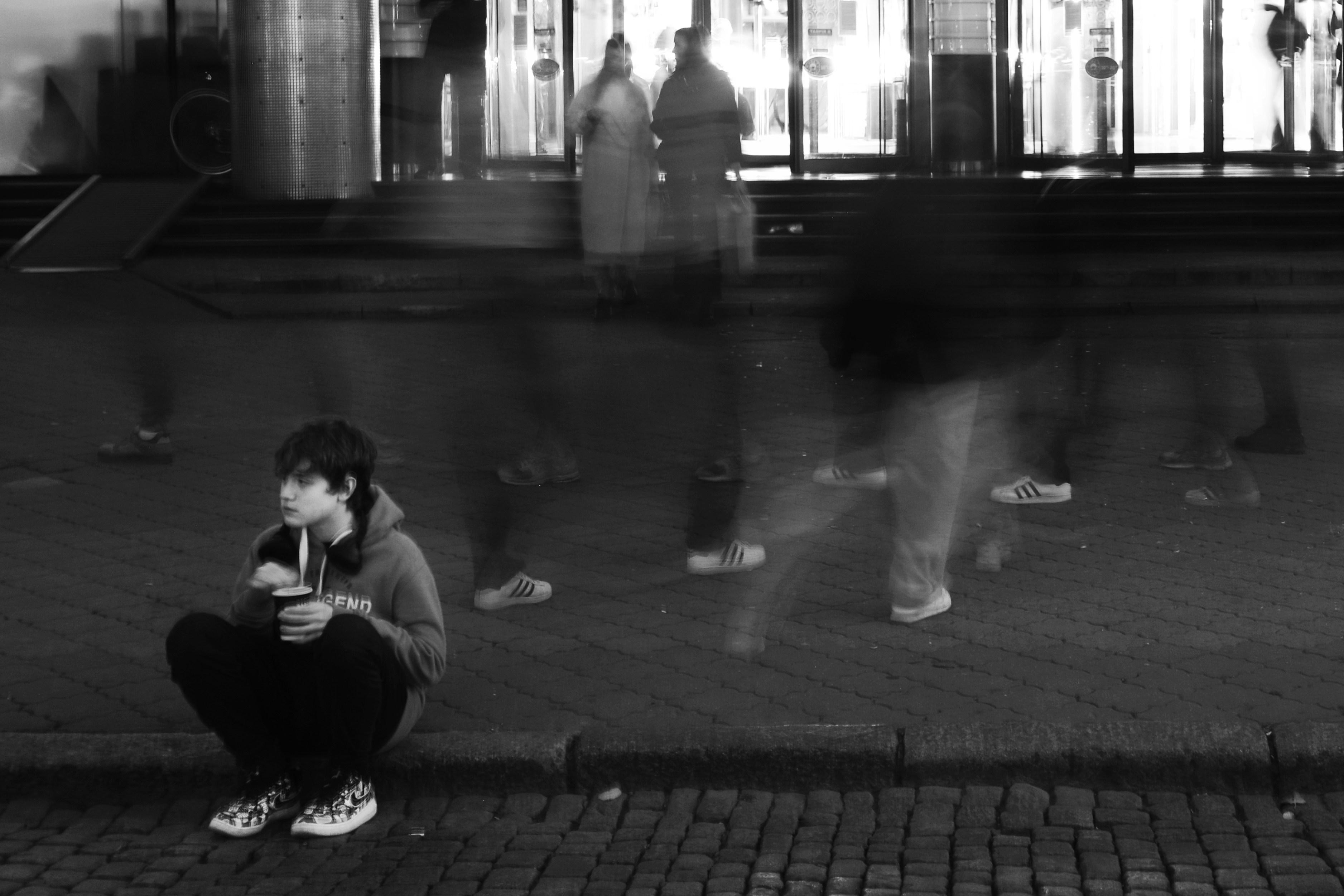Person sitting on a curb with a drink as blurred figures pass by.