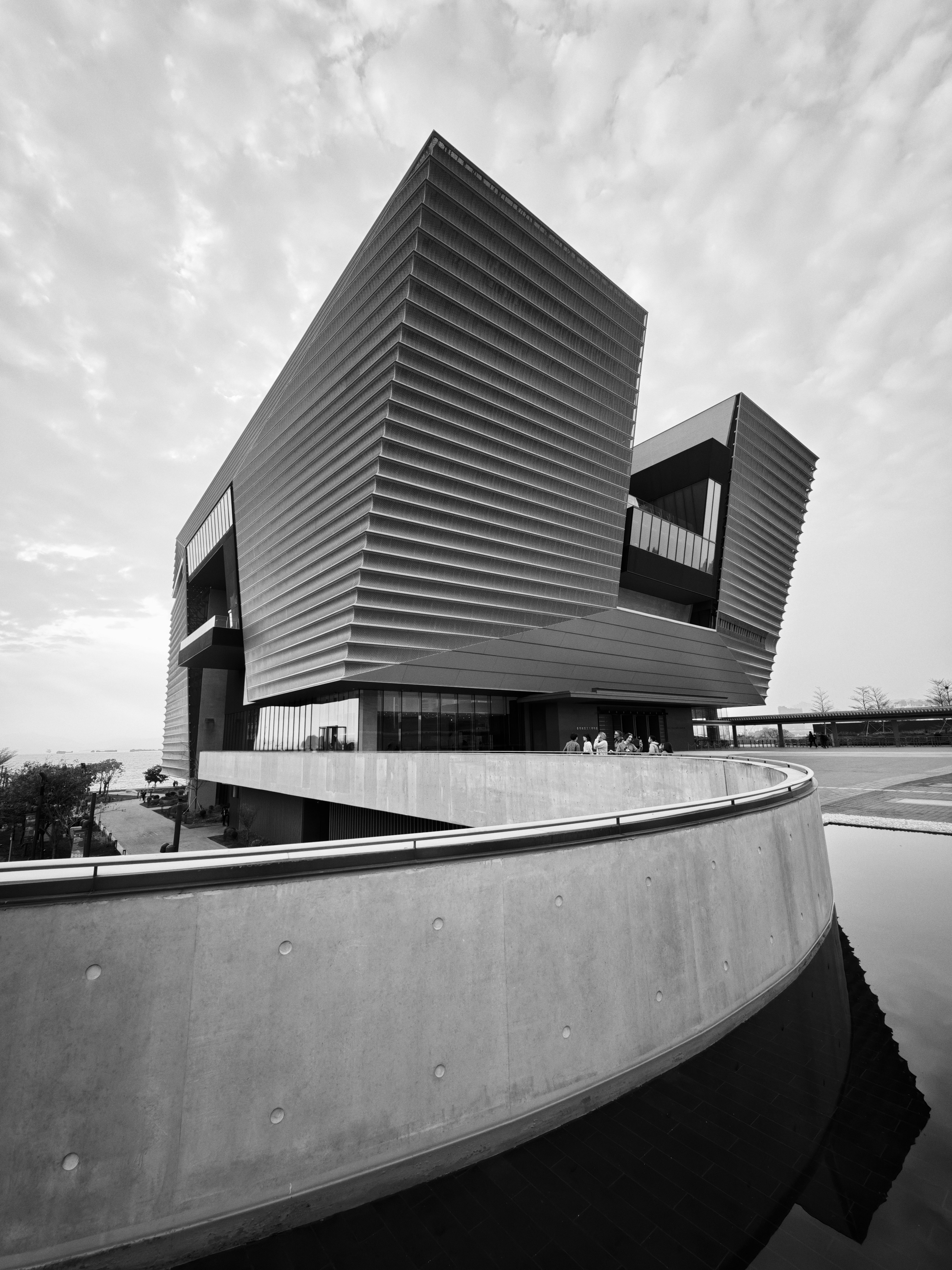 Monolithic modernist building with striped, angular volumes rises over a curved concrete barrier; black-and-white architectural study emphasizing geometry and perspective.