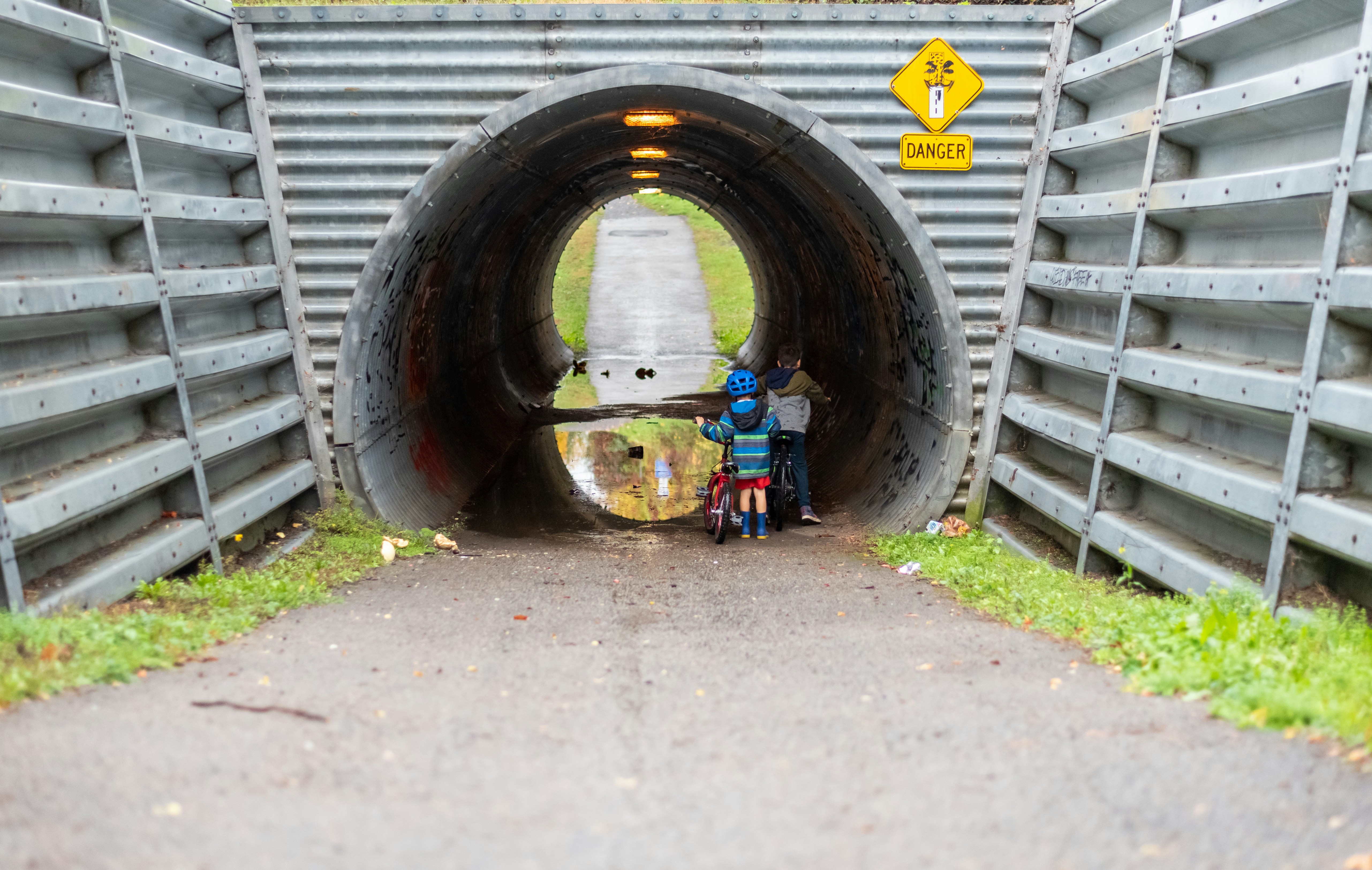 Children with bikes entering a corrugated metal tunnel, flanked by greenery, under a cloudy sky.