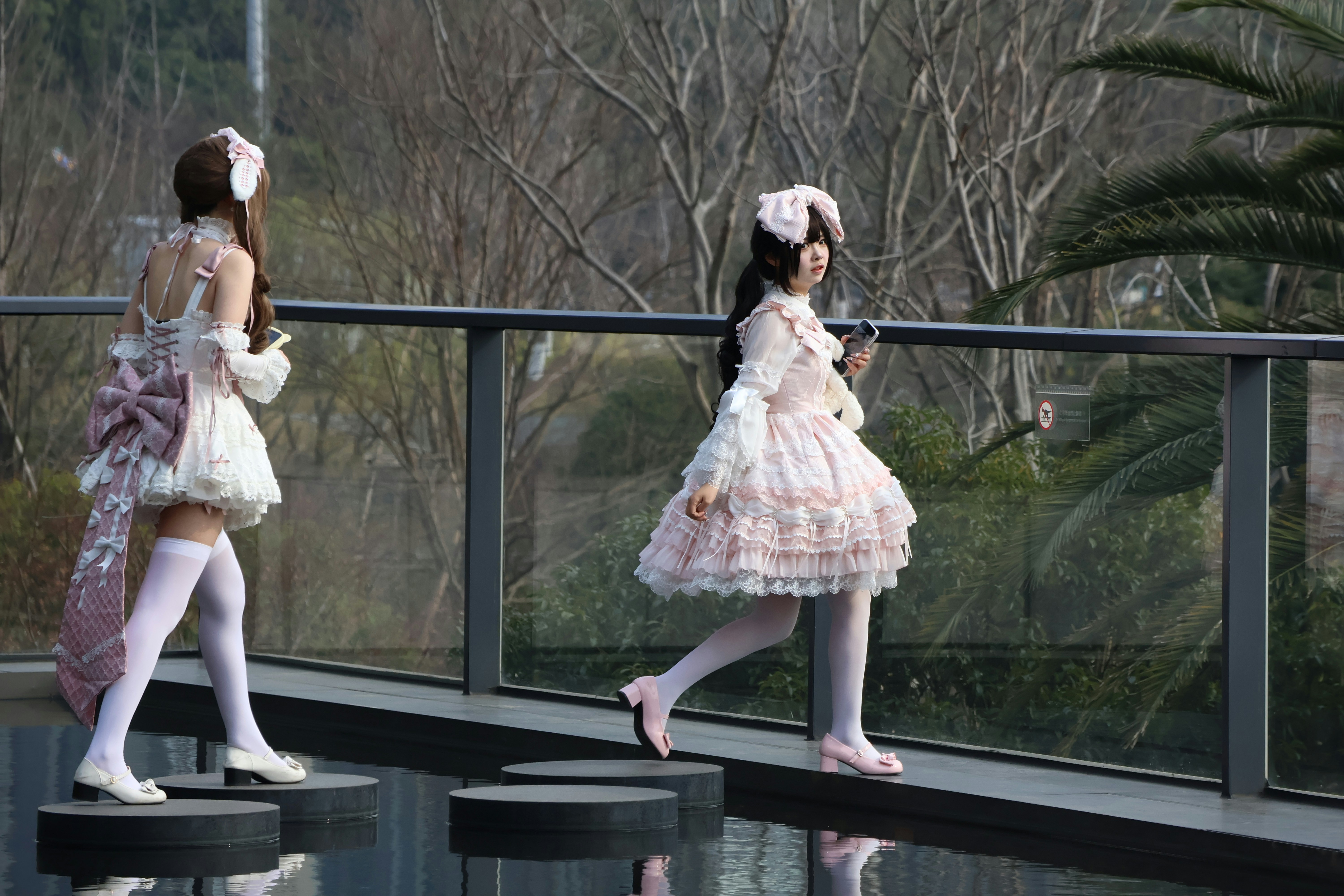 Two individuals in elaborate pink dresses stroll along a reflective walkway with a glass railing and trees in the background.