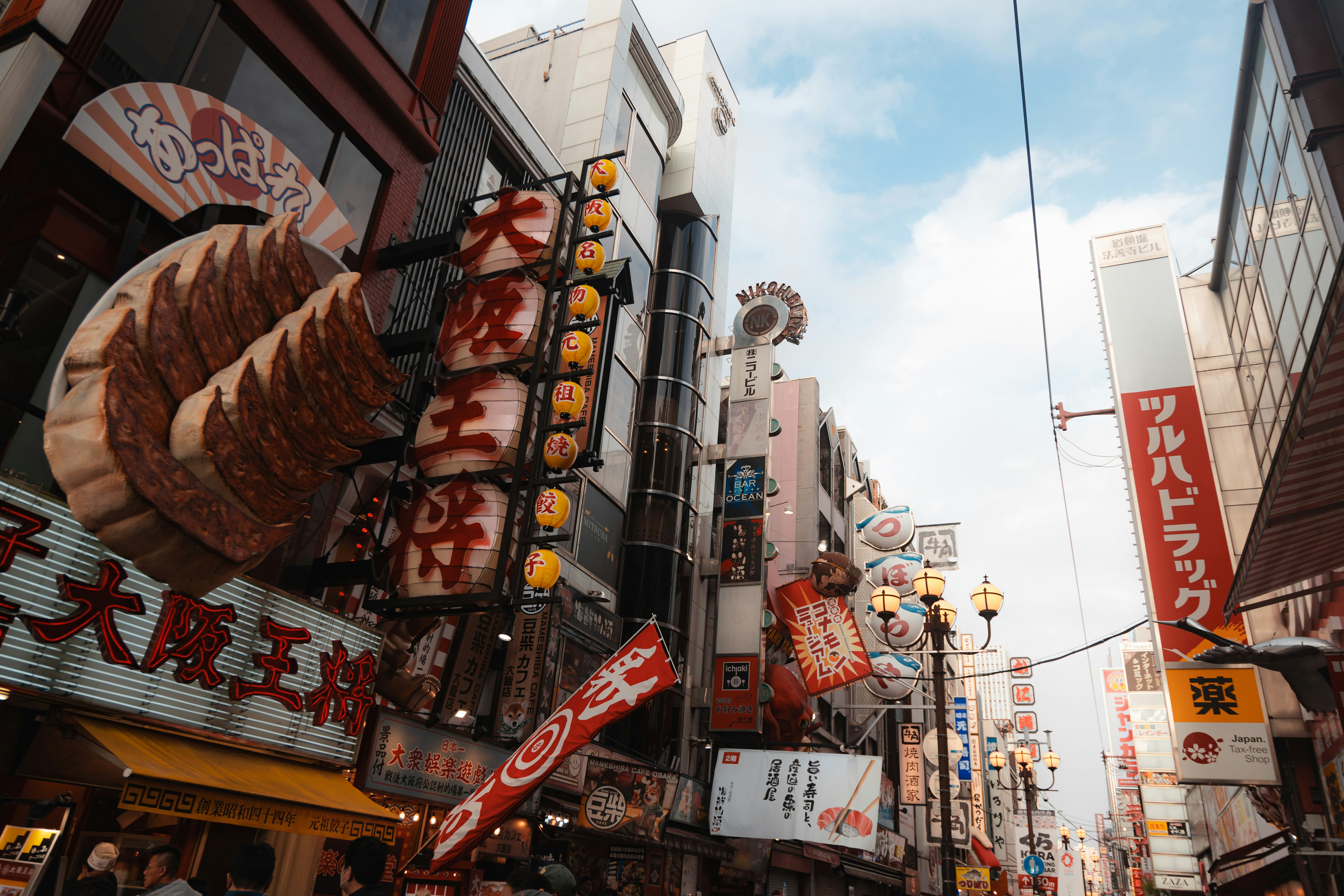Colorful shop signs and billboards line a bustling street under a bright sky in Dotonbori, Osaka.