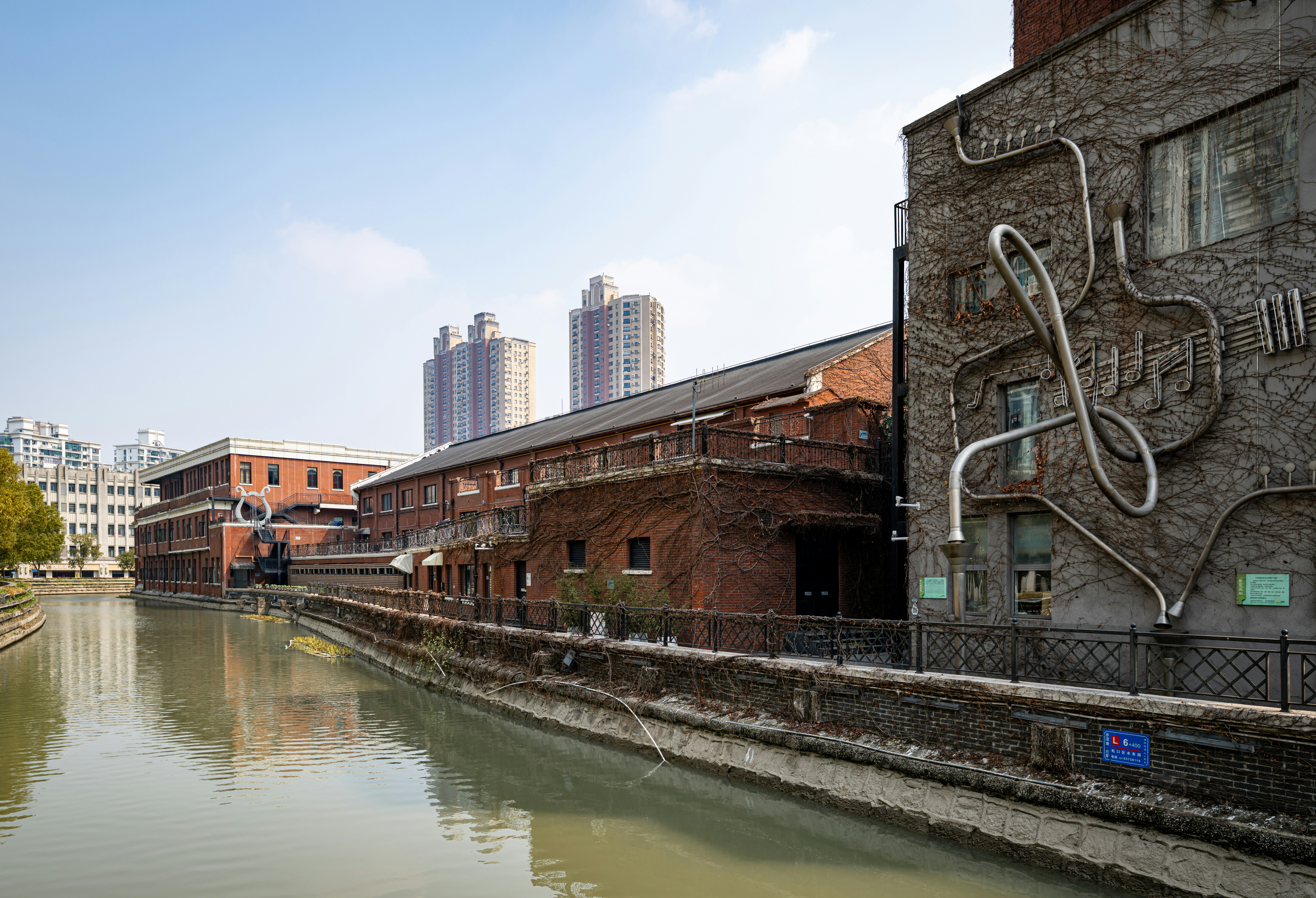 Old brick buildings adorned with metal art line a tranquil canal under a clear sky.