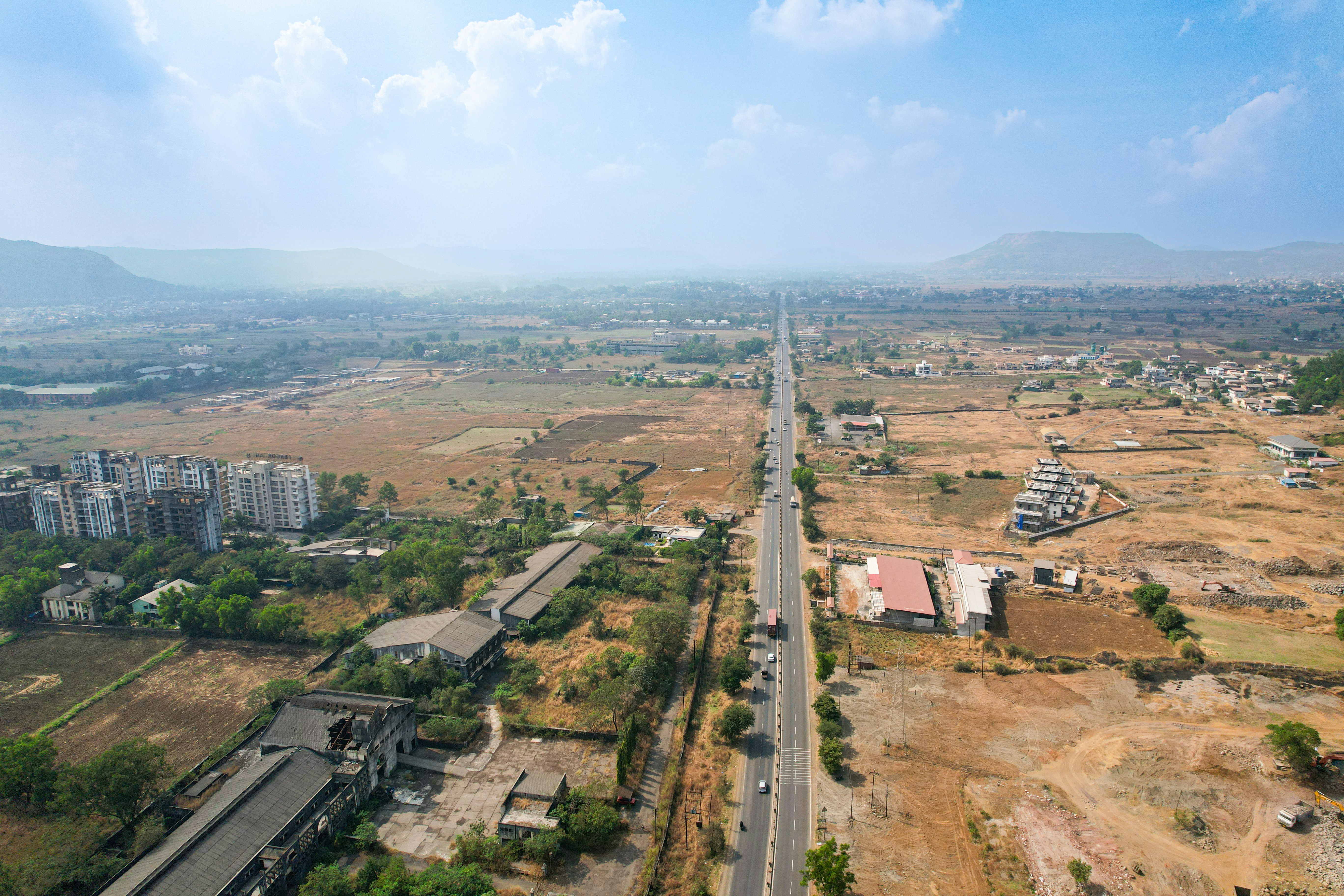 Road stretches through a blend of urban buildings and rural fields under a blue sky.