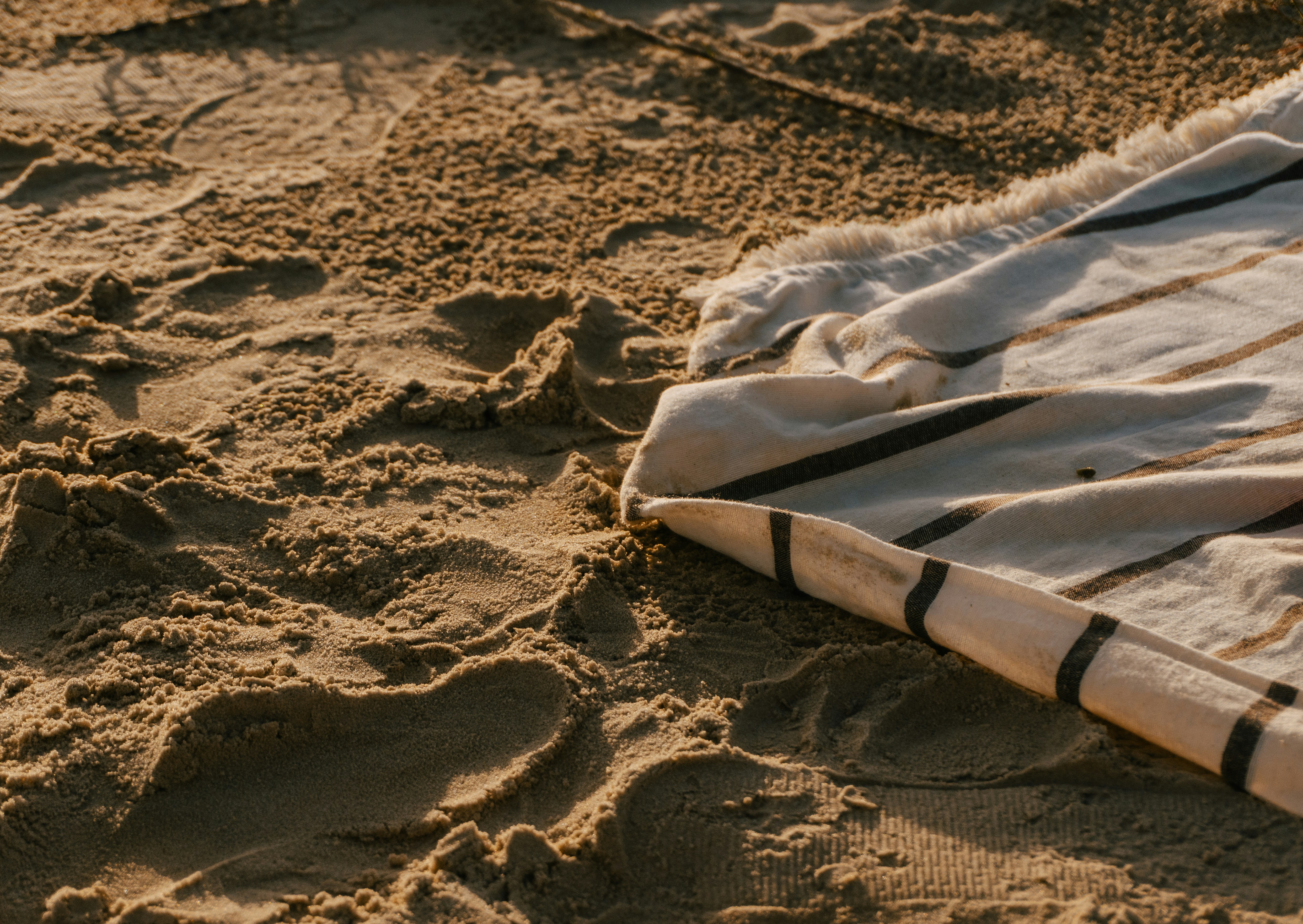 Striped blanket lies on textured beach sand under warm sunlight.