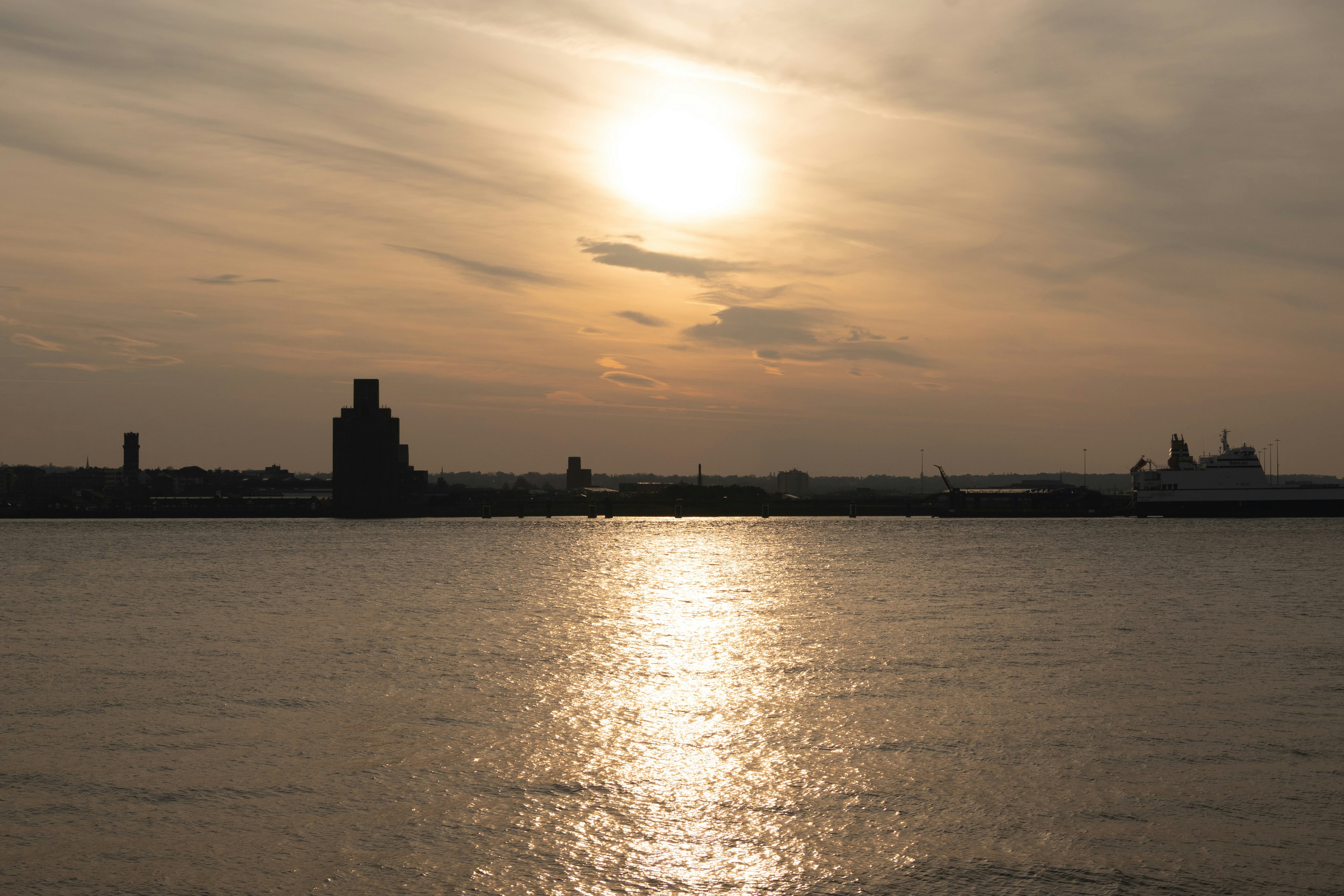 Silhouette of buildings and a ferry against an orange-hued sunset reflecting on calm waters.