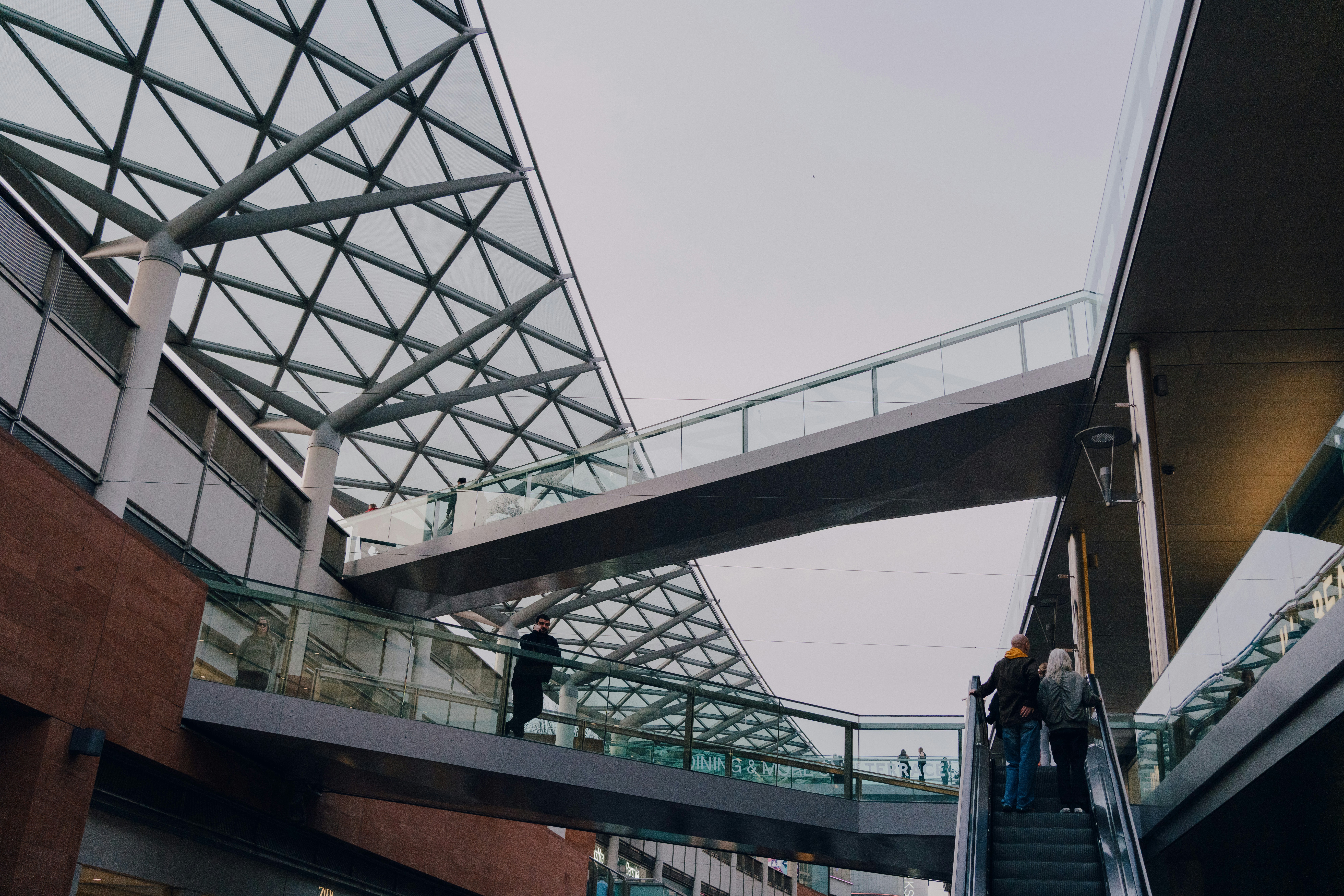 Glass walkways crisscross under a geometric glass roof in a contemporary mall.