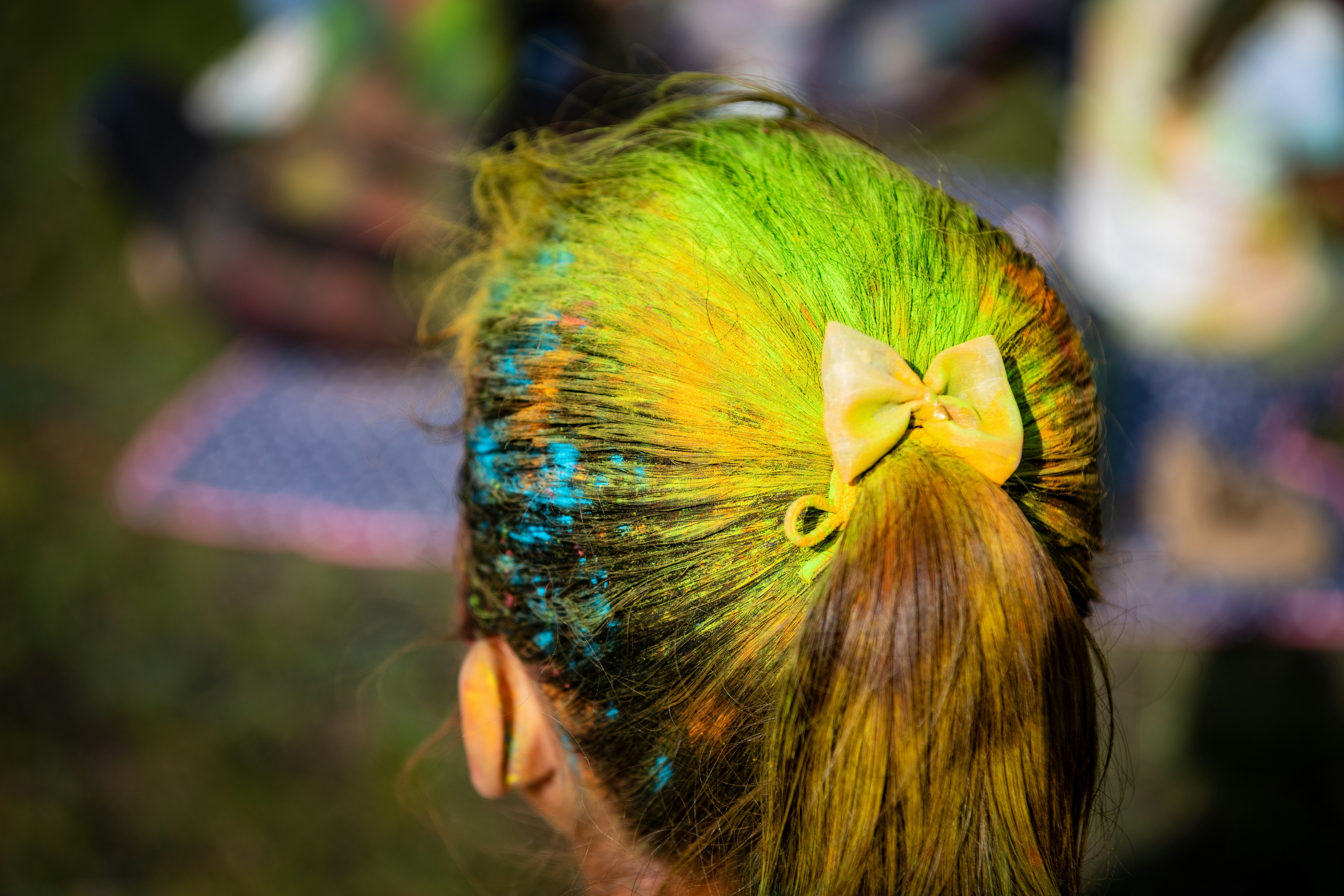 Hair adorned with vivid green, yellow, and blue colors, tied with a bow, after a Holi celebration.