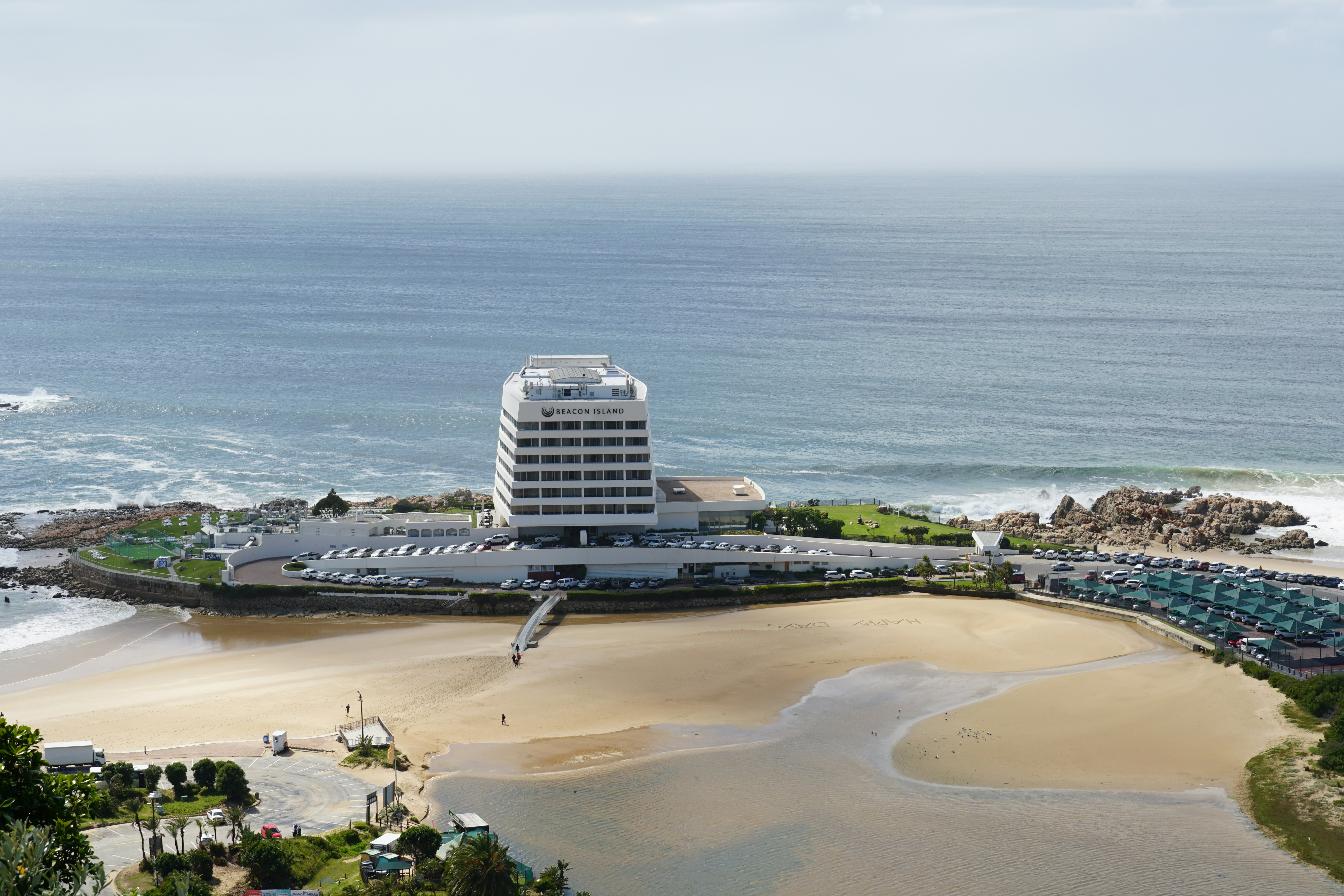 Modern hotel by the oceanfront with surrounding sandy beach and rocky shore.