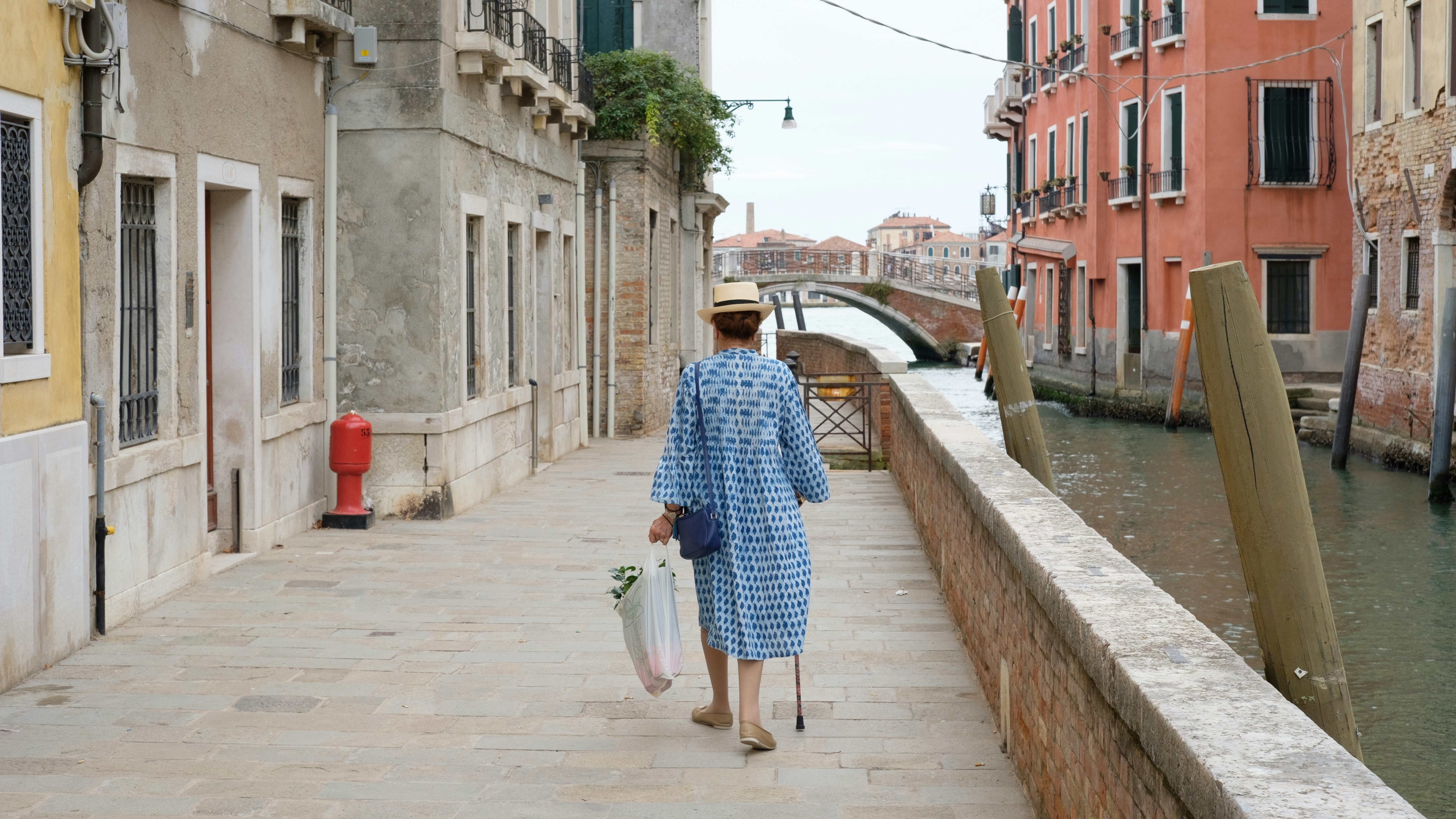 Woman in a blue dress walks beside a Venetian canal past colorful buildings and a small bridge.