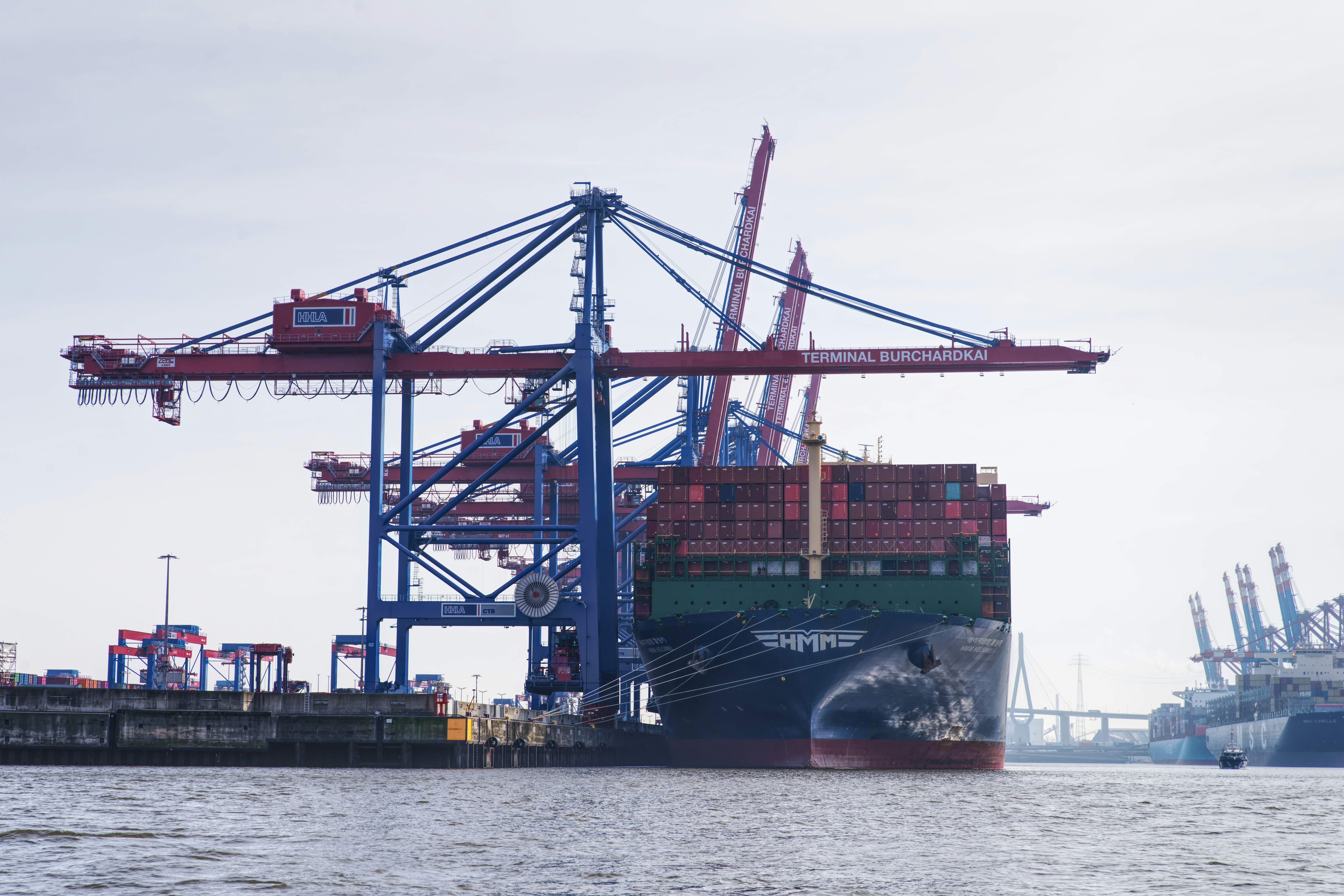 Large cargo ship docked at a port, surrounded by towering cranes under a cloudy sky.