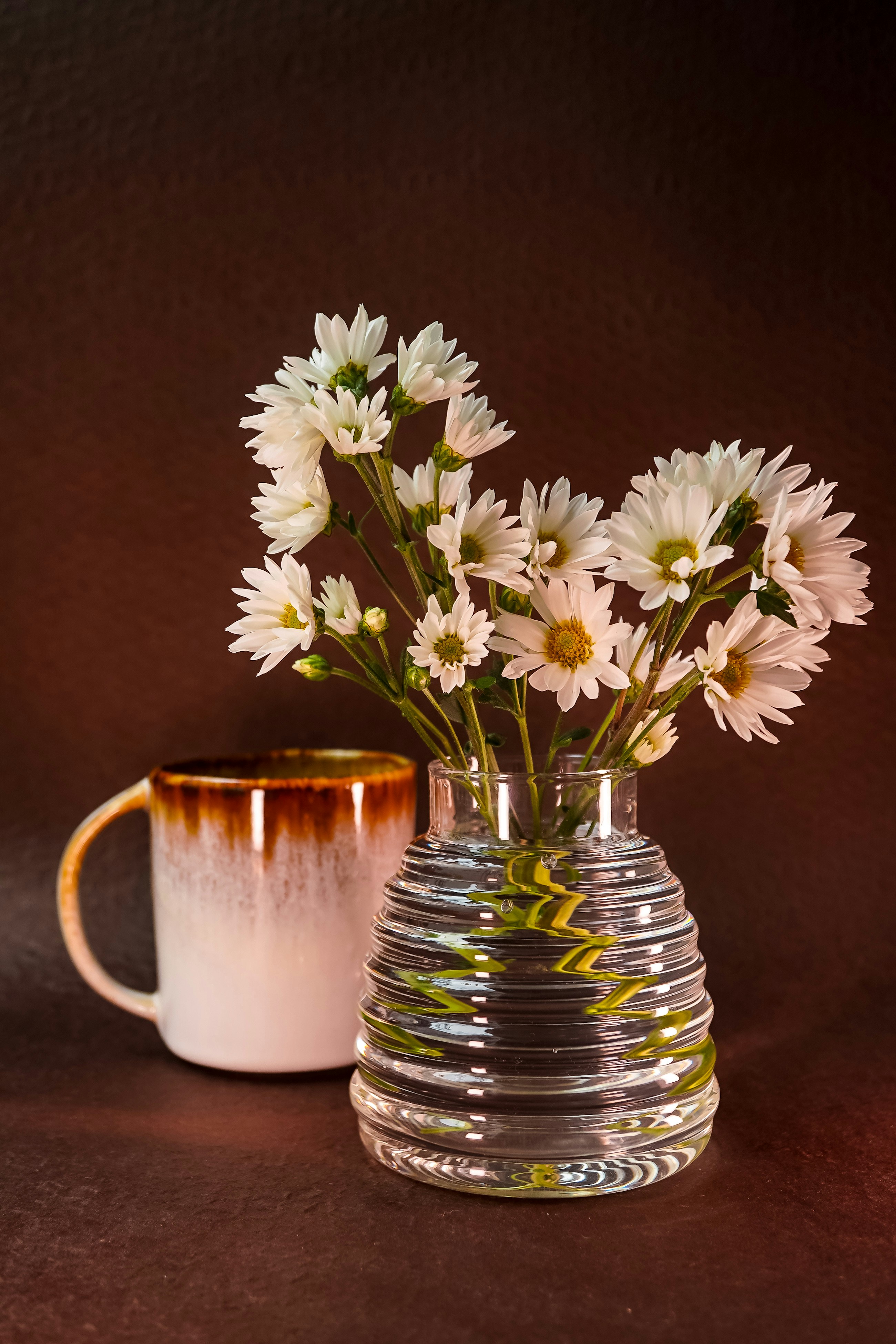 White daisies in a ribbed glass vase beside a ceramic mug with a brown gradient on a dark background.