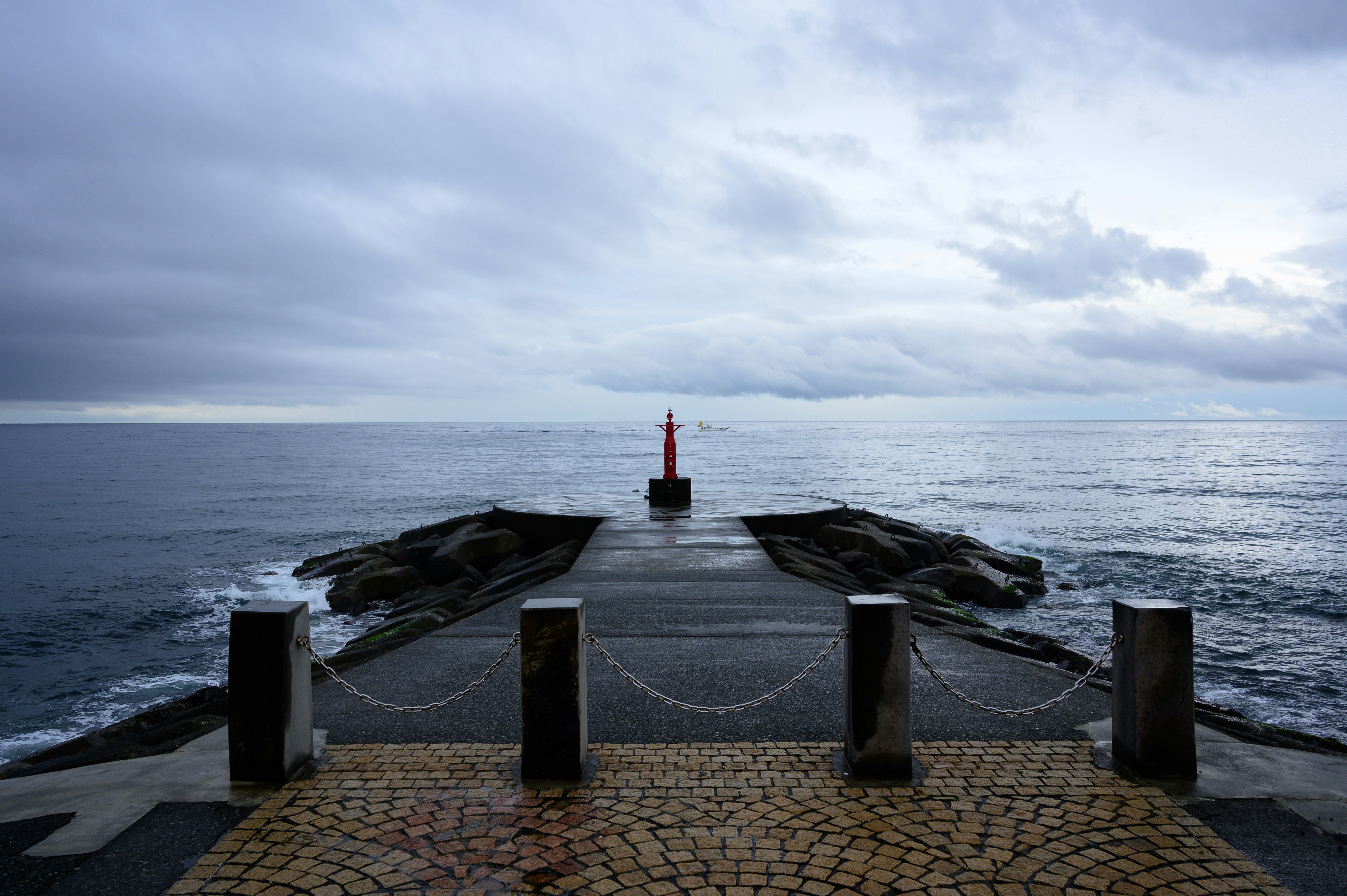 Stone pier leading to a red lighthouse under overcast skies.