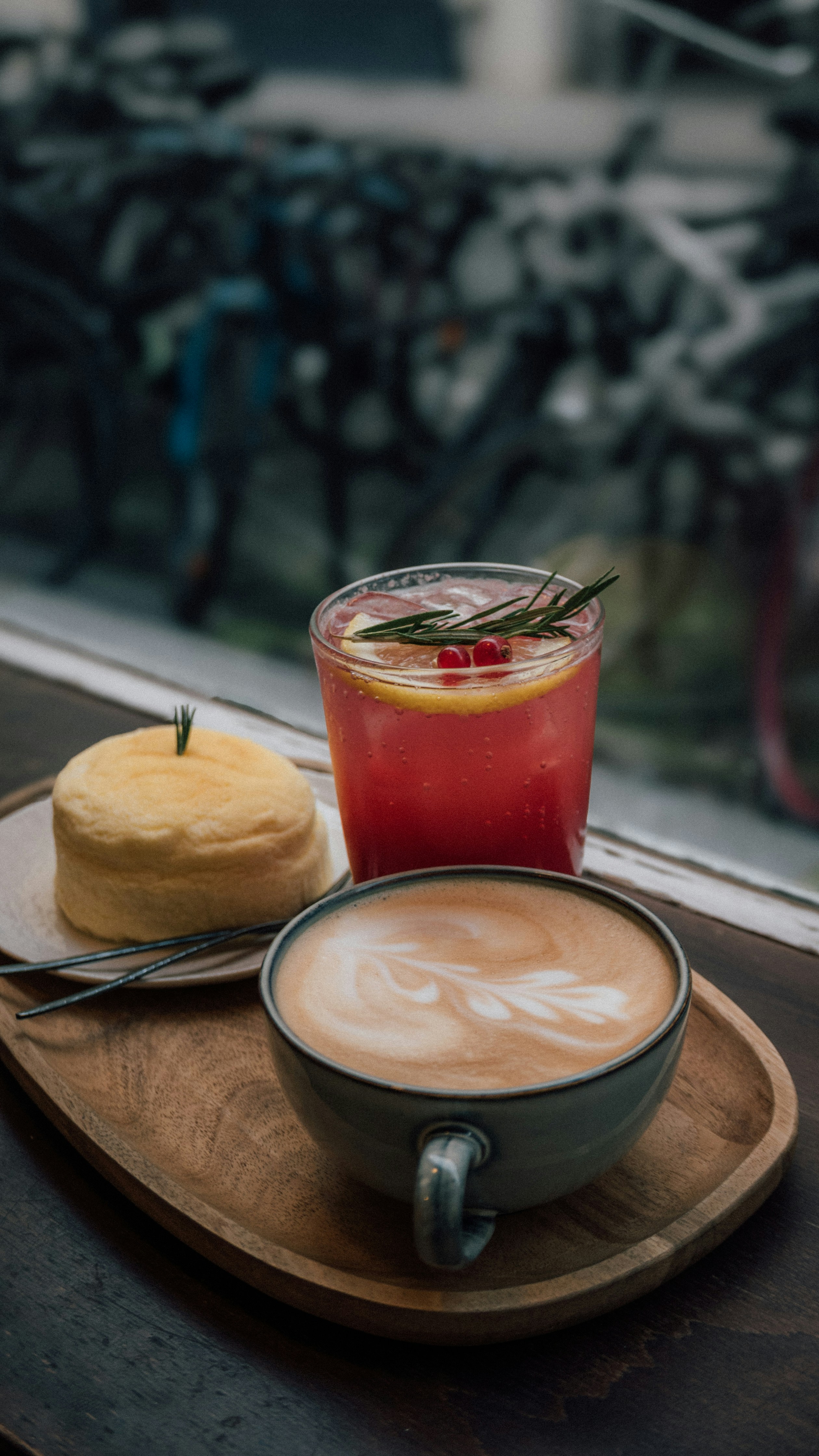 A frothy cappuccino with latte art sits on a wooden tray beside a round pastry and a pink citrus beverage on a window-side table.