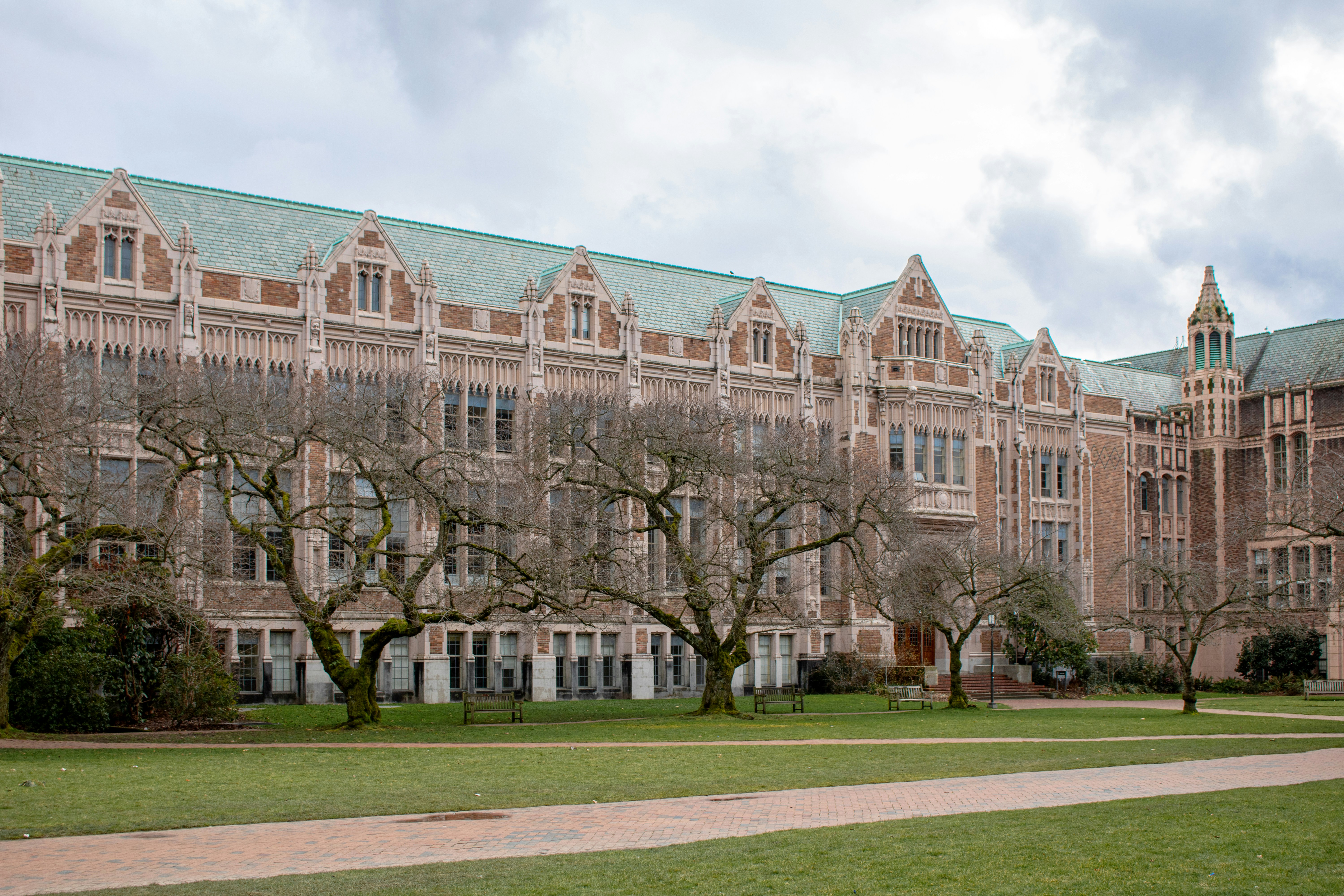 This wide shot showcases Smith Hall, a prominent building on the University of Washington campus completed in 1938. Its construction marked the completion of the Liberal Arts Quadrangle. The hall, with its blend of stone and brick, features numerous windows and gabled roofs characteristic of Gothic Revival architecture. A row of bare-branched trees lines the front, contrasting with the green lawn and paved pathway. The overcast sky casts a diffused light. Smith Hall houses the departments of Geography, History, Law, Societies & Justice, and Political Science.