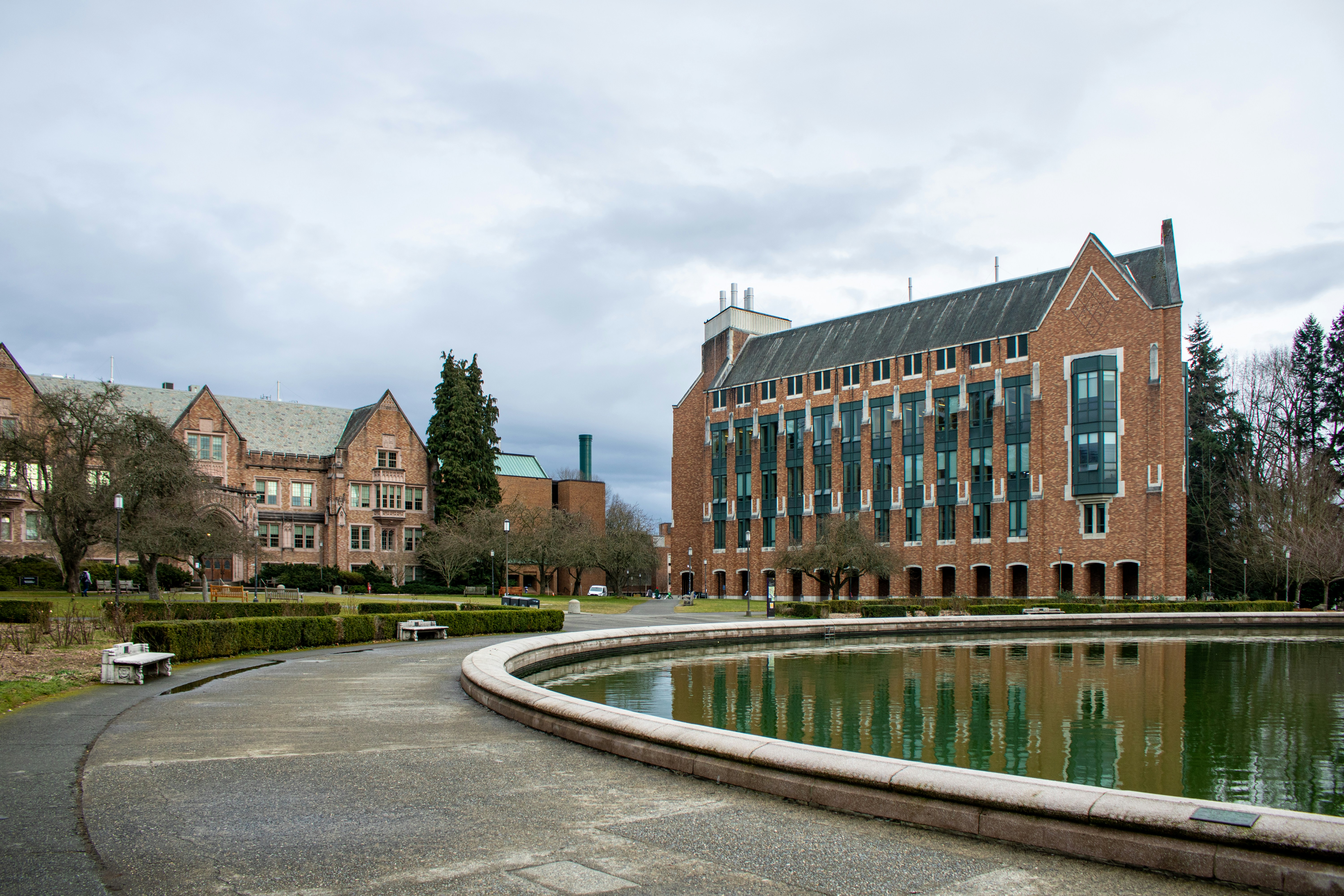 Brick university buildings reflected in a circular fountain under a cloudy sky.