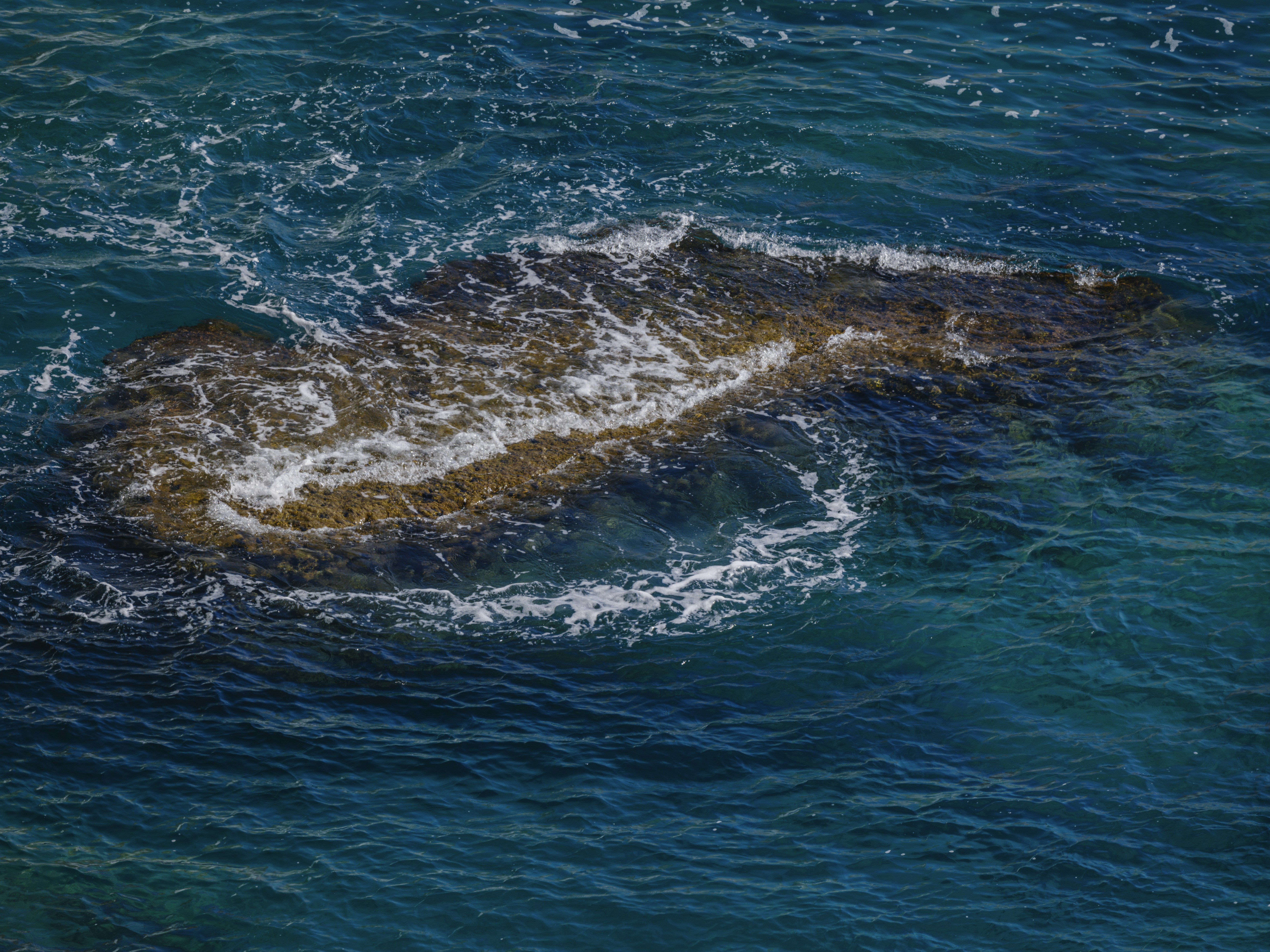 Waves swirl around a solitary rocky outcrop in vibrant blue waters.