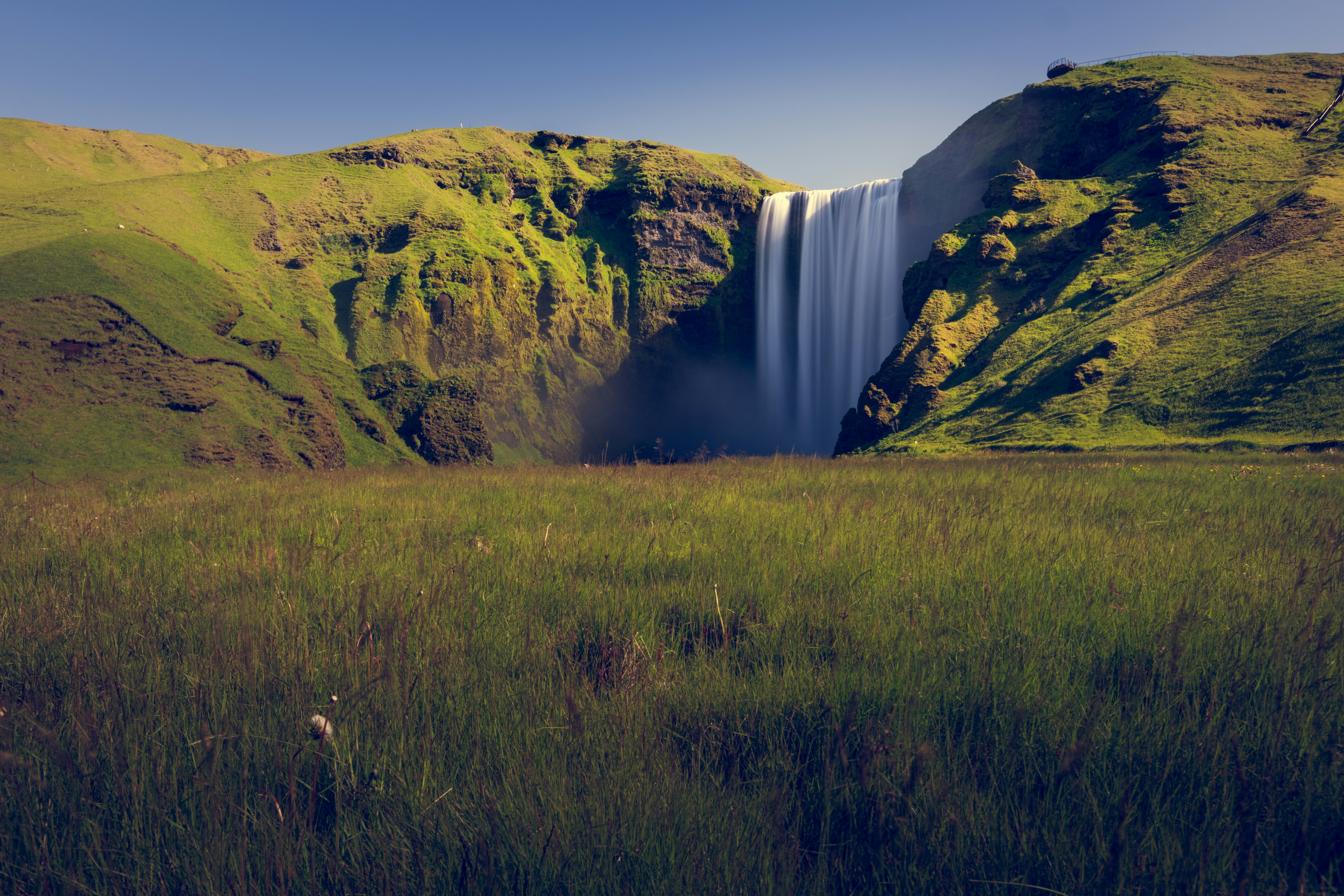 Waterfall plunges down a lush green cliff under clear blue skies.