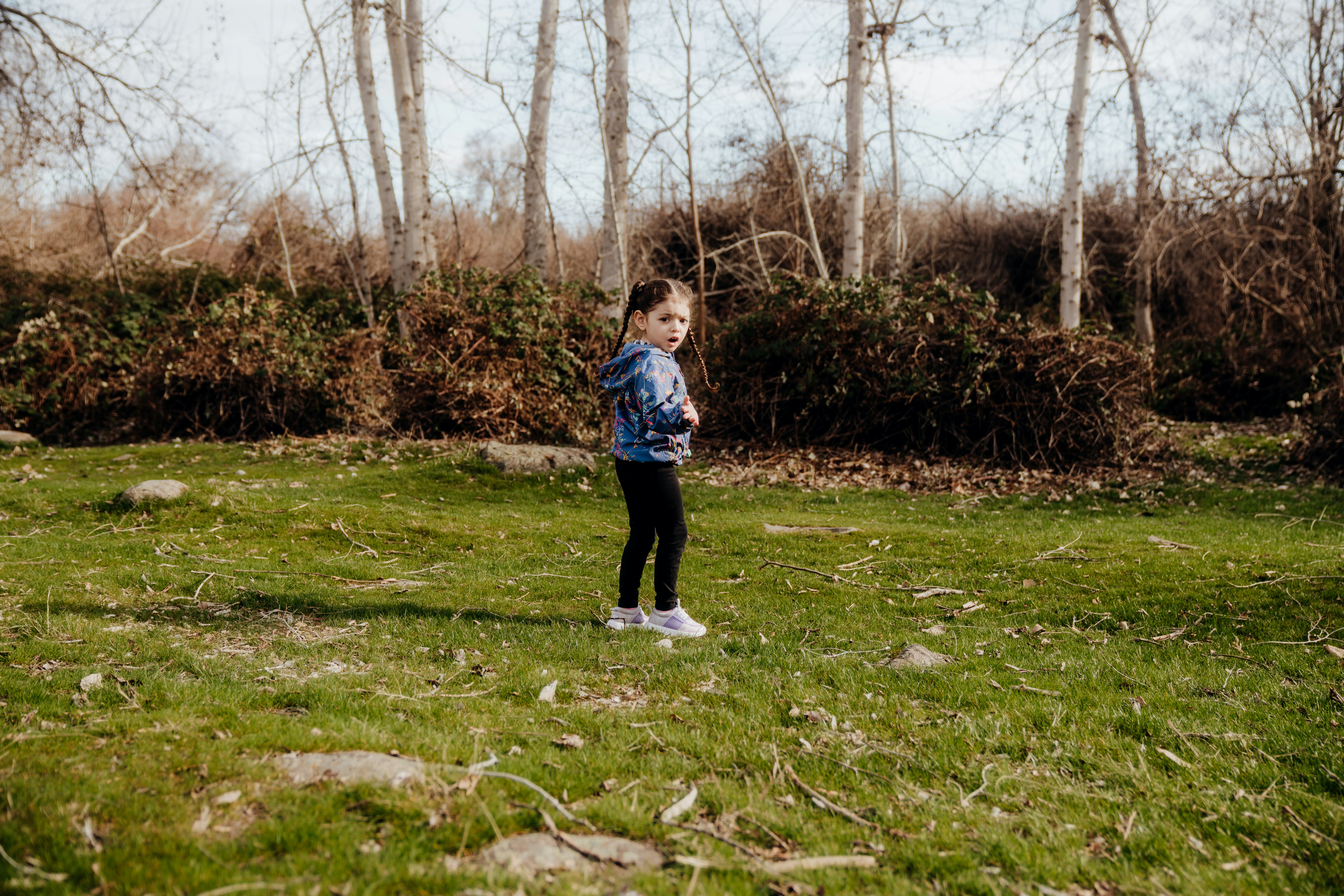Child in a vibrant hoodie stands on lush green grass near bare trees.