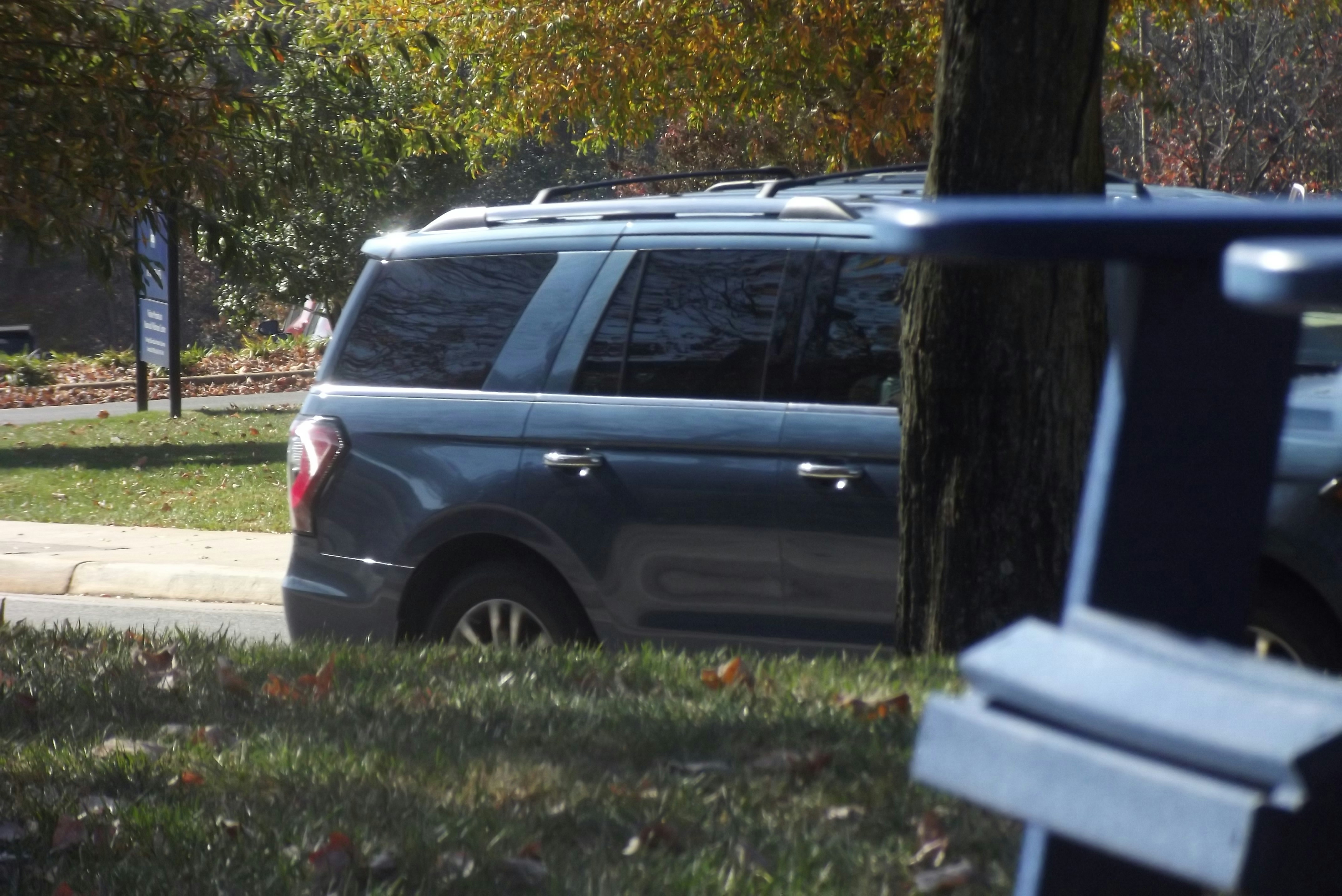 Gray SUV parked along a tree-lined park road with autumn leaves on the ground, blue bench visible in the foreground.