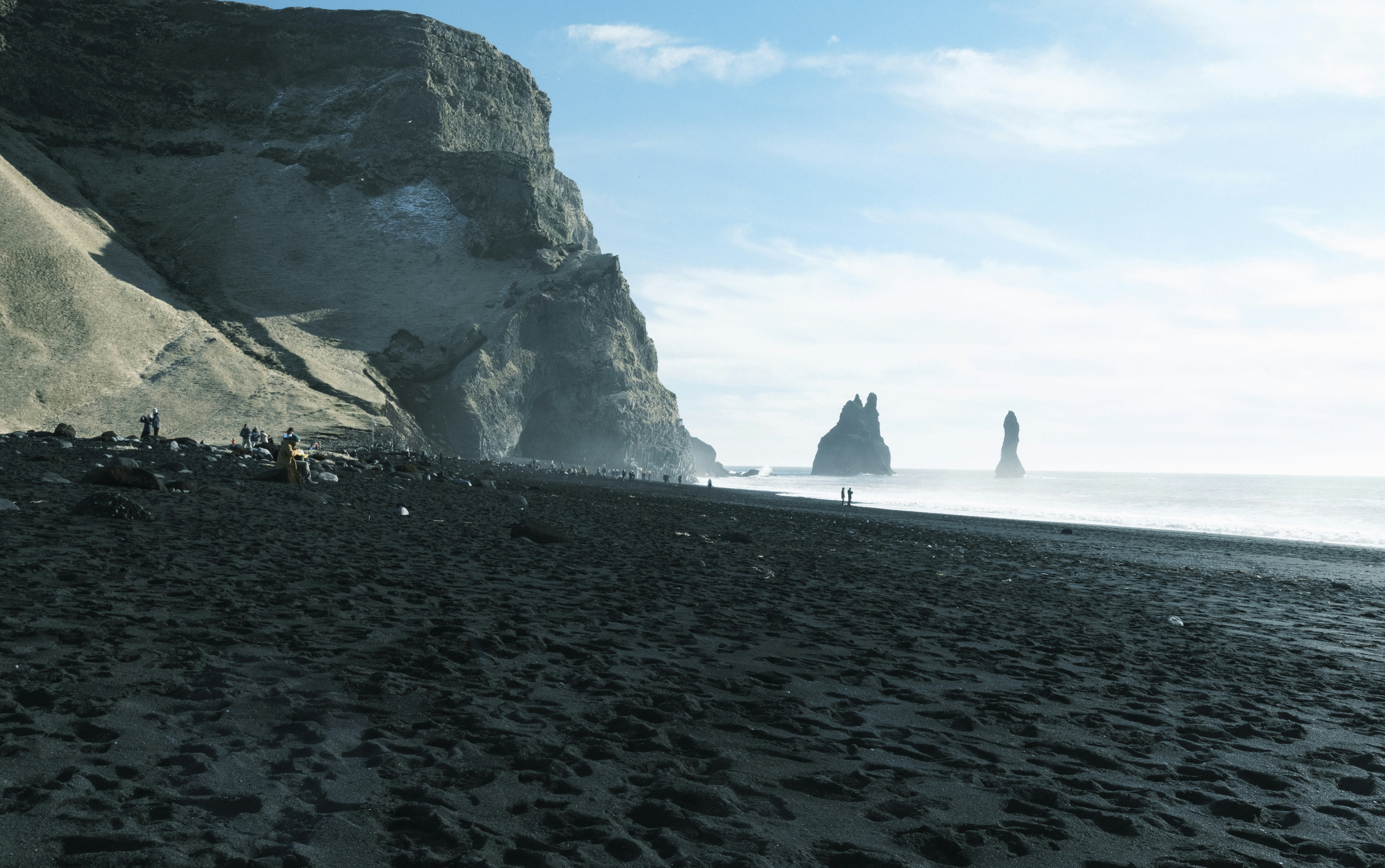 Black sand beach with towering sea stacks and rugged cliffs under a bright sky.