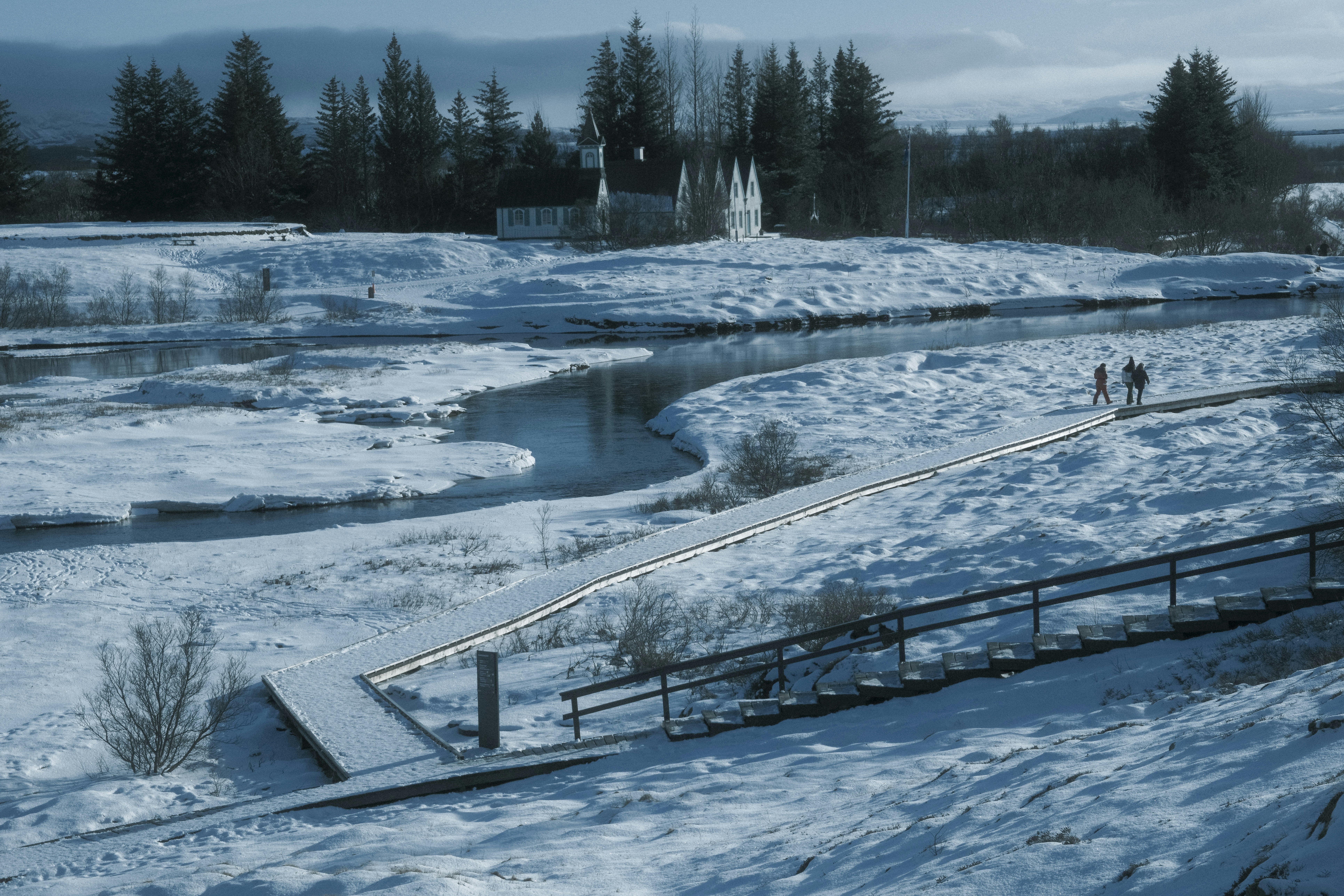 Snow-covered landscape with a boardwalk and river, flanked by trees and distant houses.