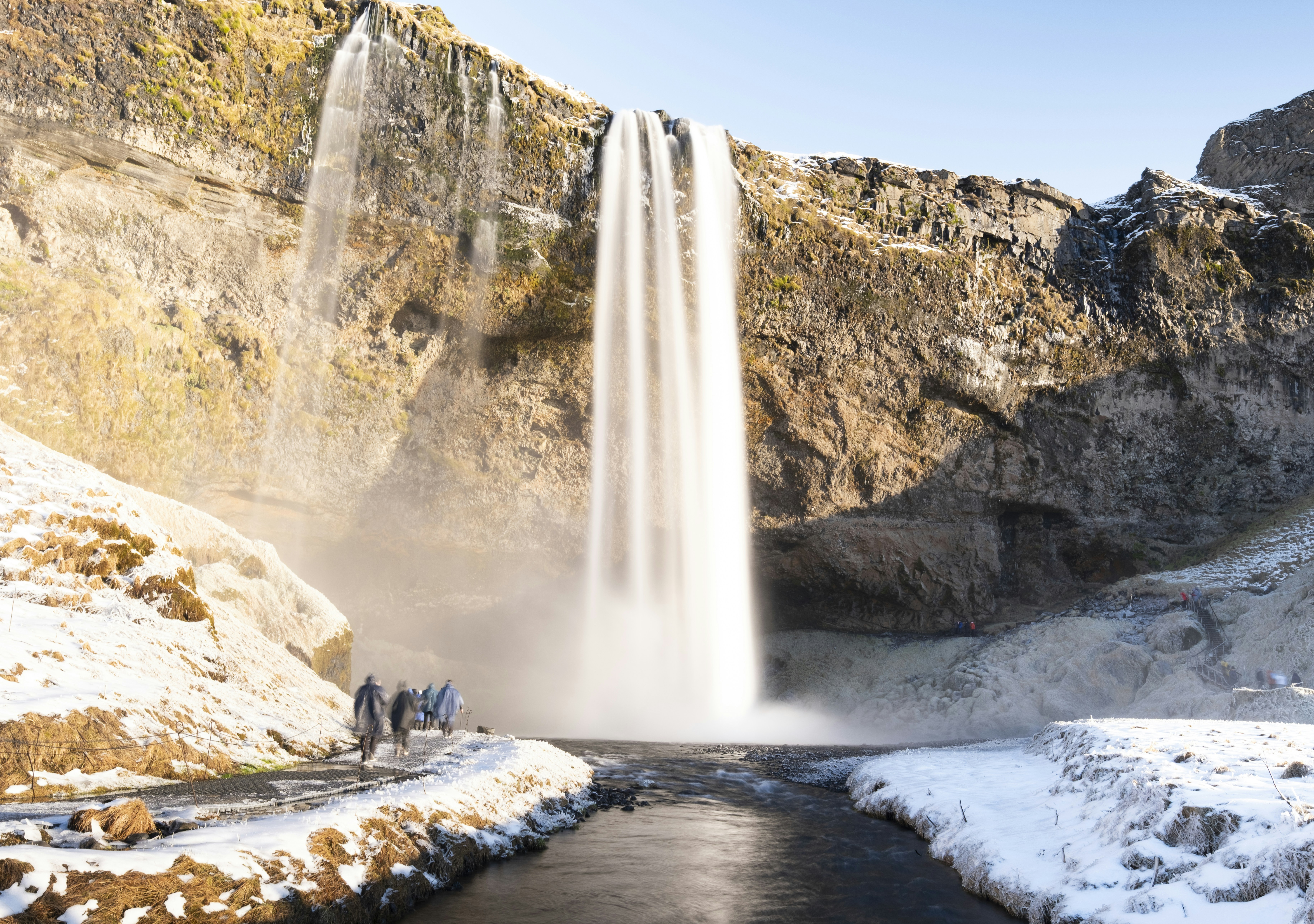 Waterfall cascades over rocky cliffs surrounded by snow, with visitors walking on a snowy path.