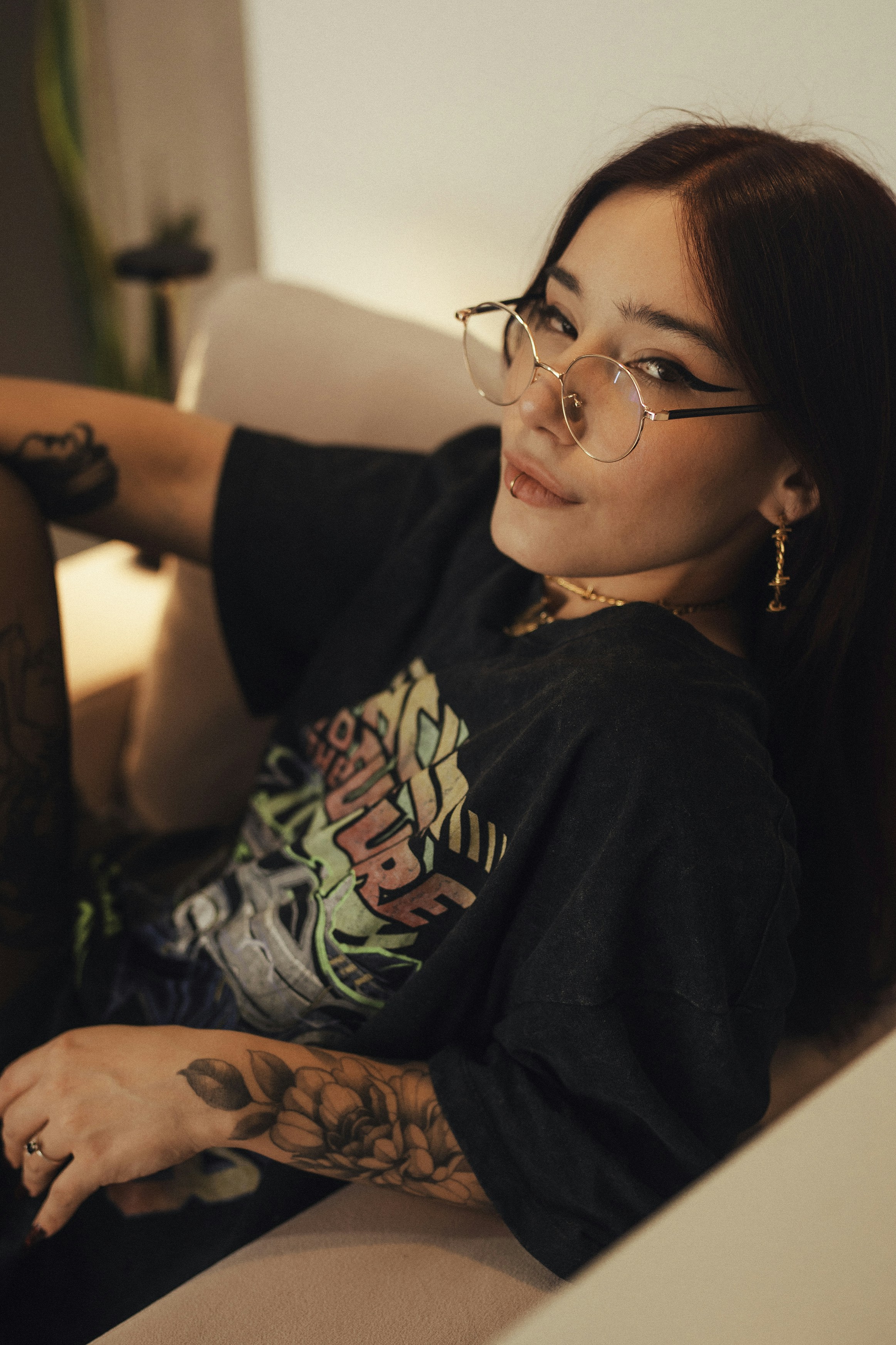 Young woman lounging casually with oversized graphic tee and stylish glasses, showcasing intricate tattoos on her arms.