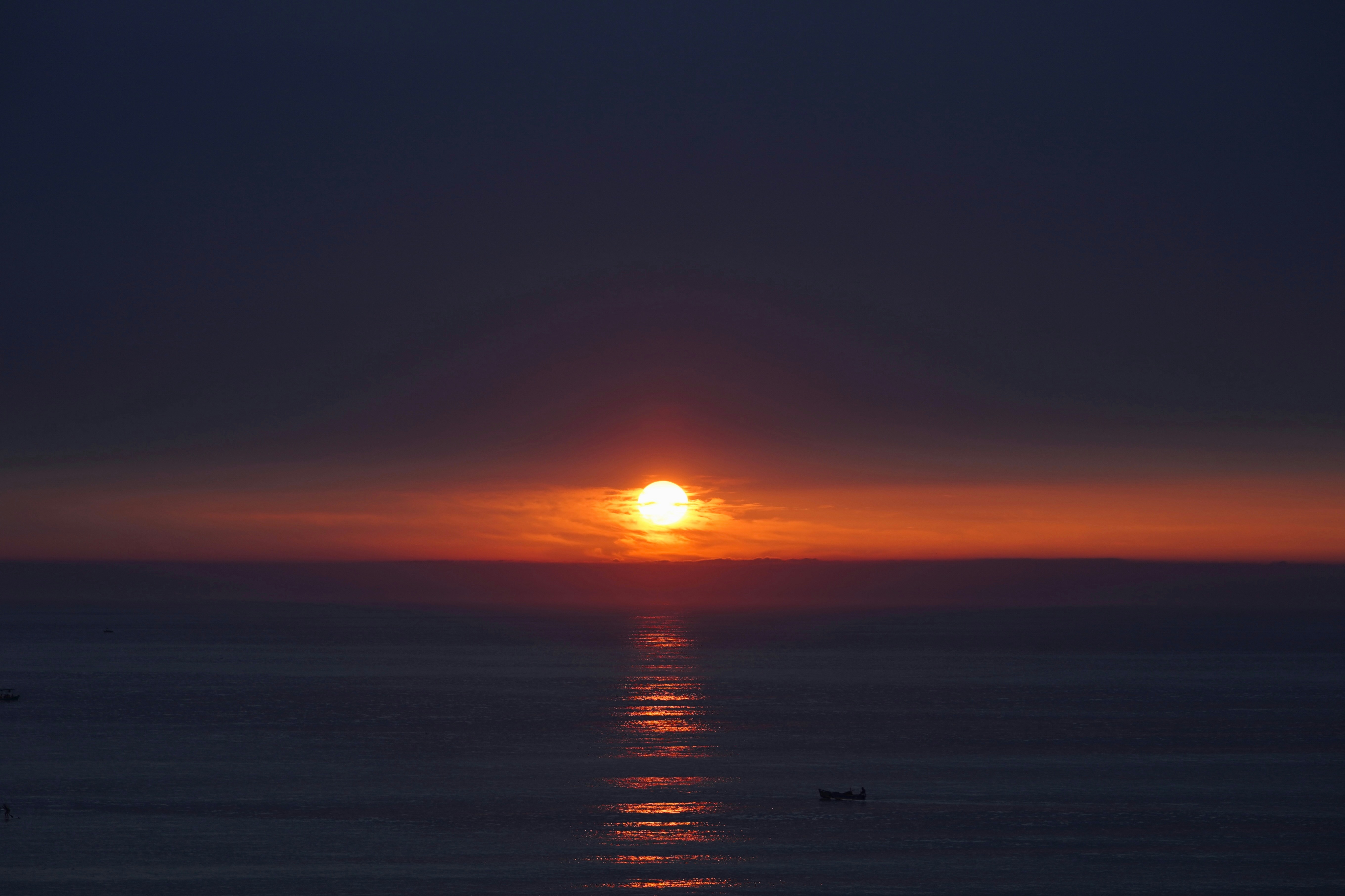 Sunset casts a fiery glow over the ocean, silhouetting a distant boat.