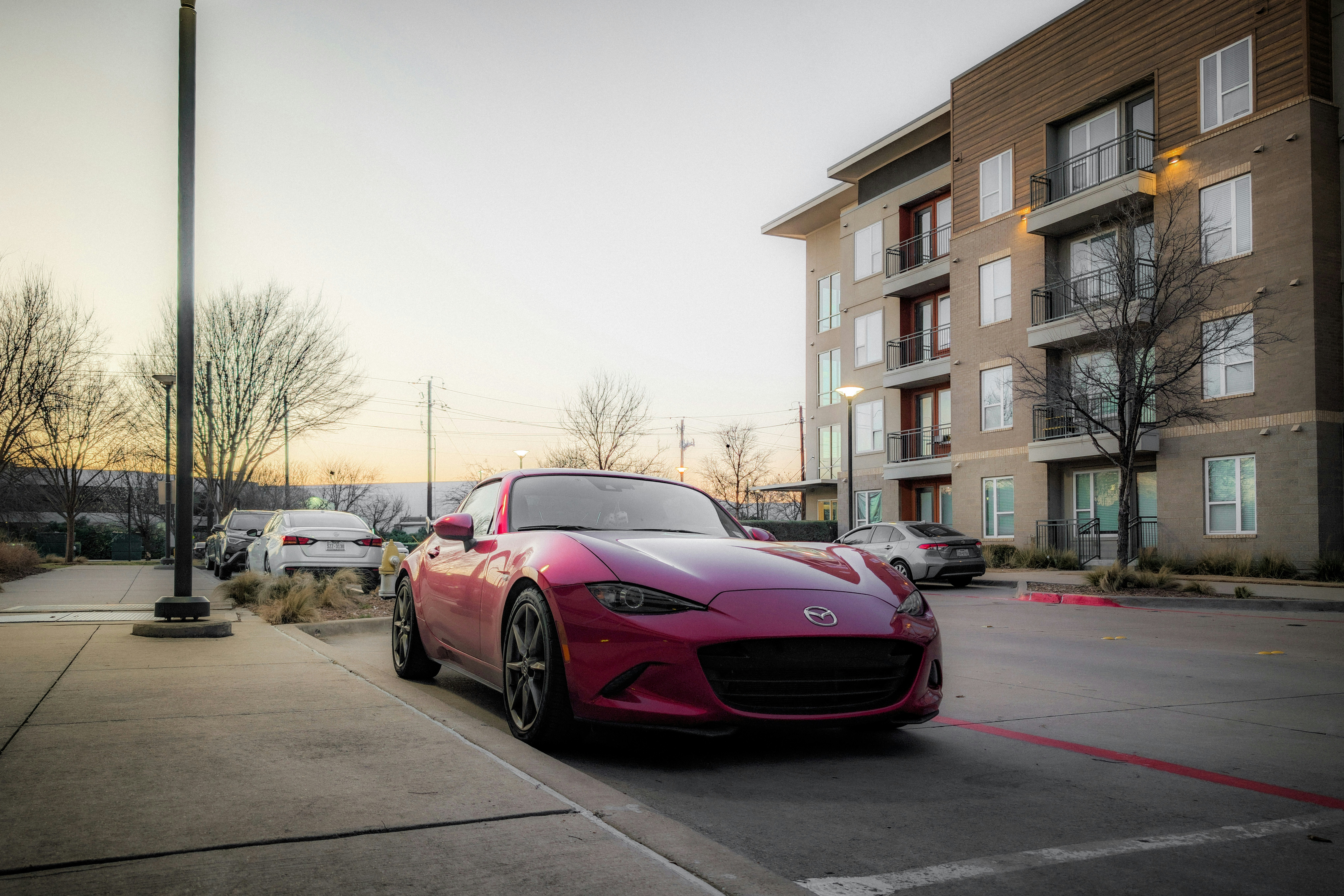 Red Mazda Miata parked beside a modern apartment building at dusk.