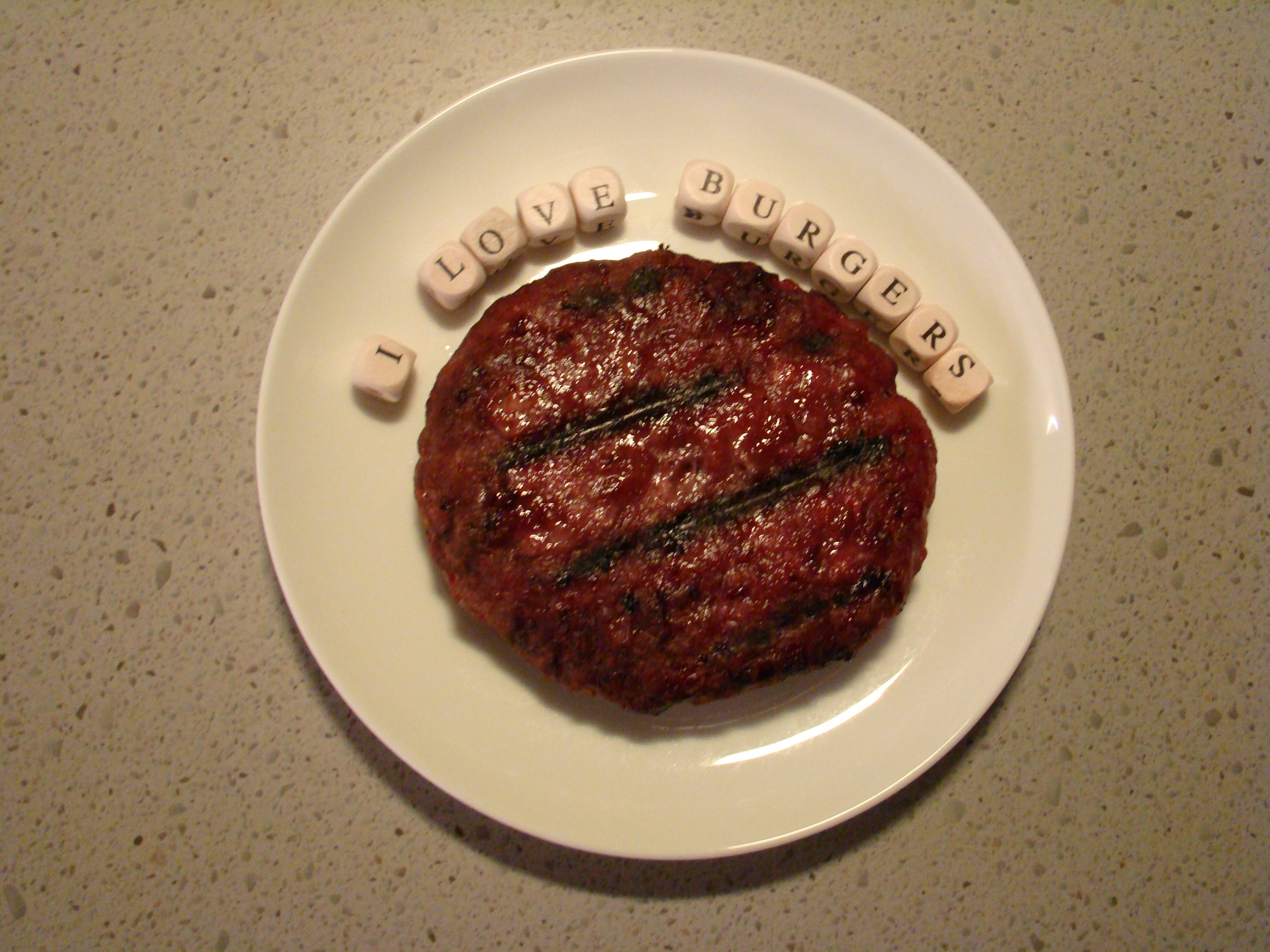 Beef burger patty with pronounced grill marks sits on a plain white plate, surrounded by Scrabble-style tiles spelling 'I LOVE BURGERS'.