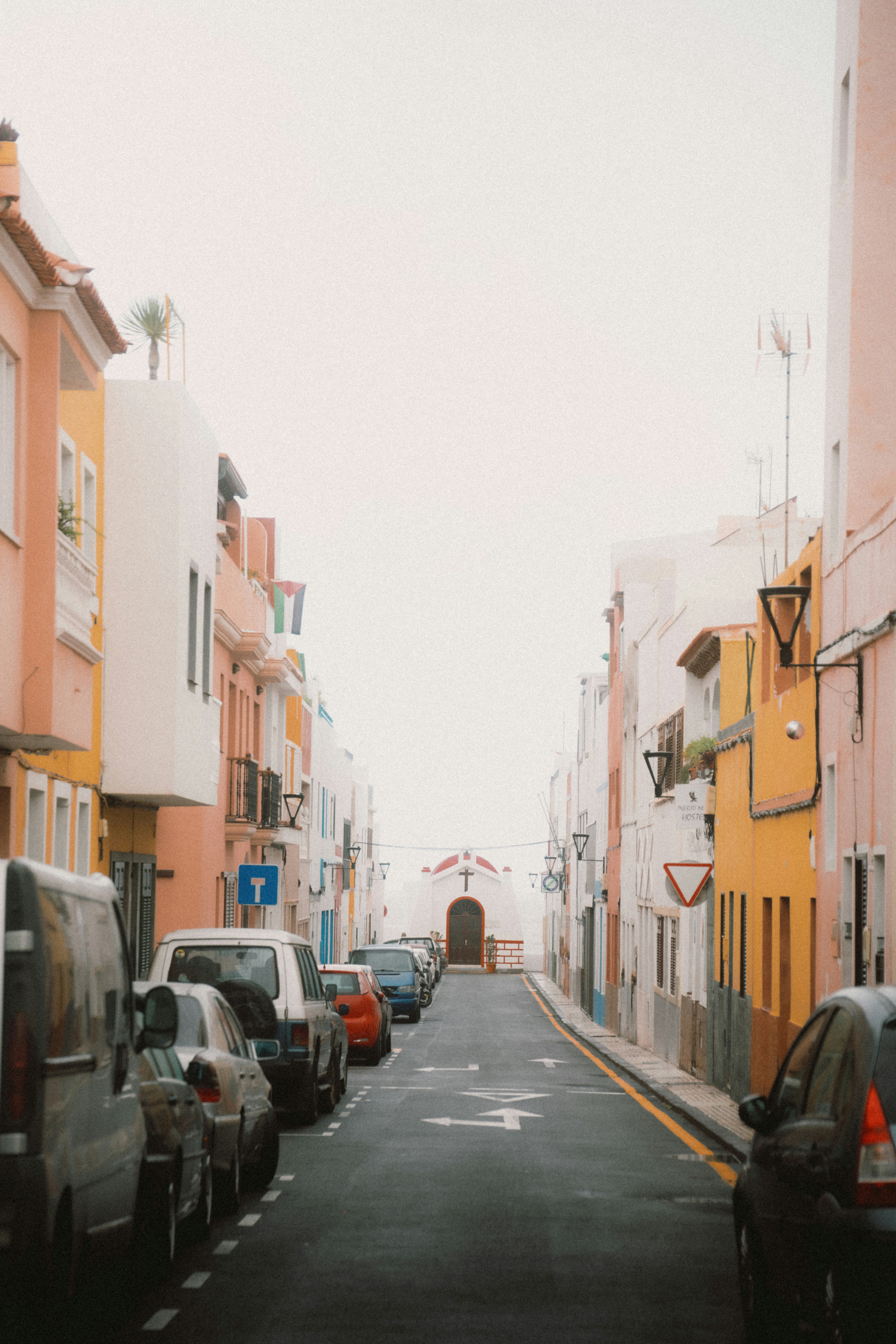 Charming street lined with pastel-colored buildings and parked cars, leading to an inviting archway at the end. The scene captures the essence of a quaint coastal town.