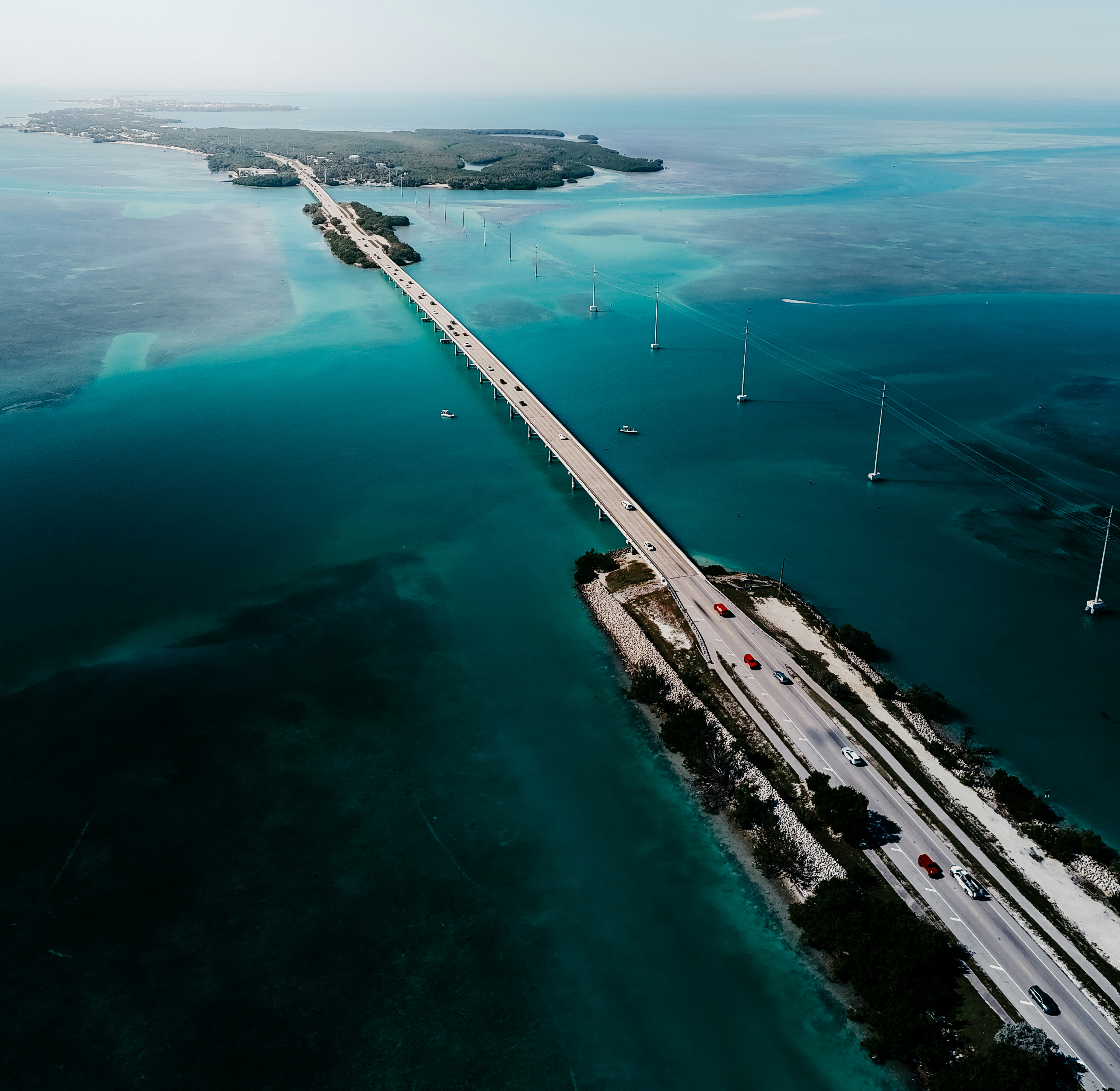 Aerial view of a winding road stretching over vibrant turquoise waters, bordered by lush greenery and dotted with boats. The scene captures the essence of coastal travel.