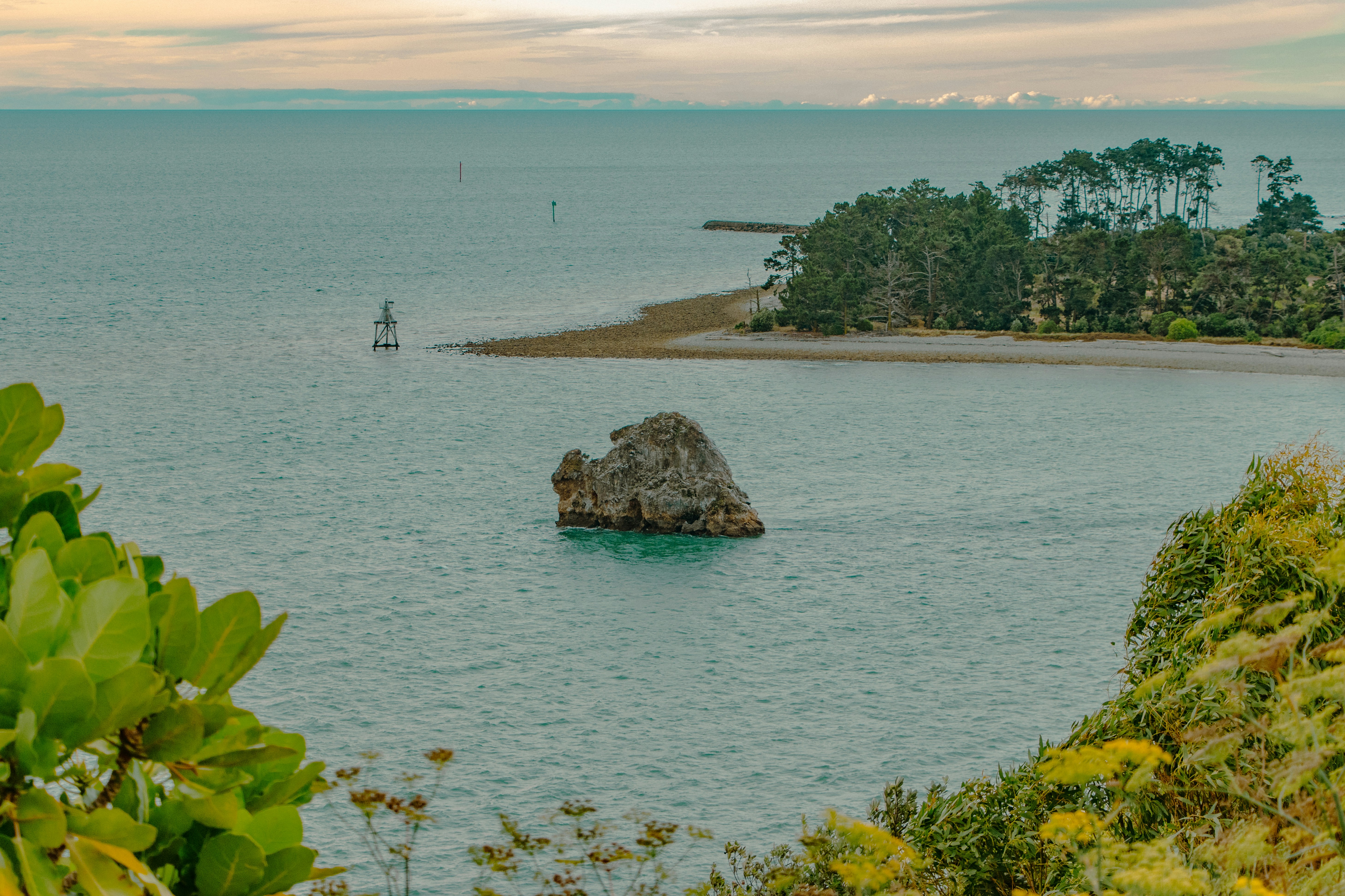 Coastal scene with a solitary rock formation surrounded by calm sea and distant trees.