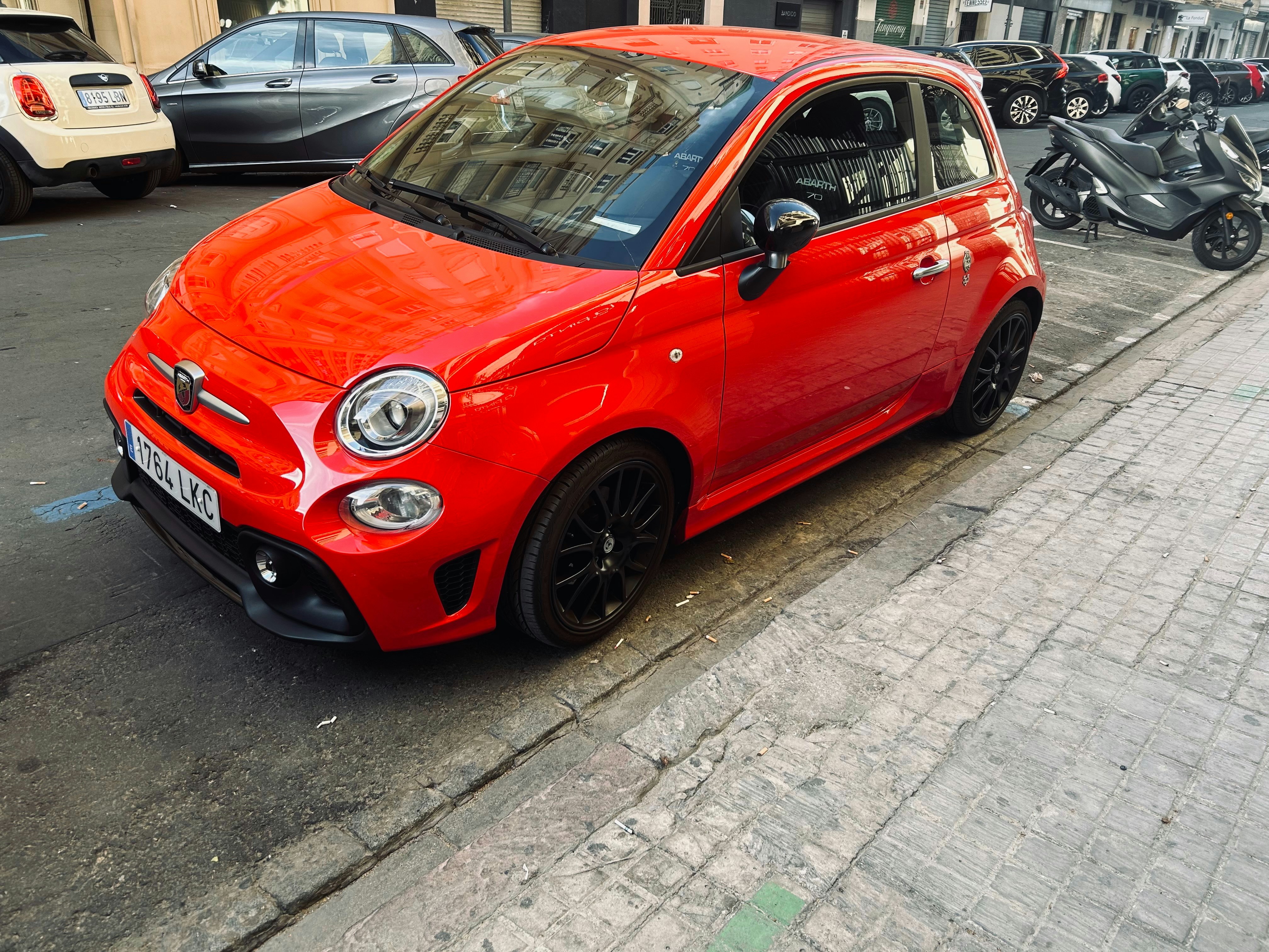 Red Abarth 595 parked on an urban street with surrounding vehicles and buildings.