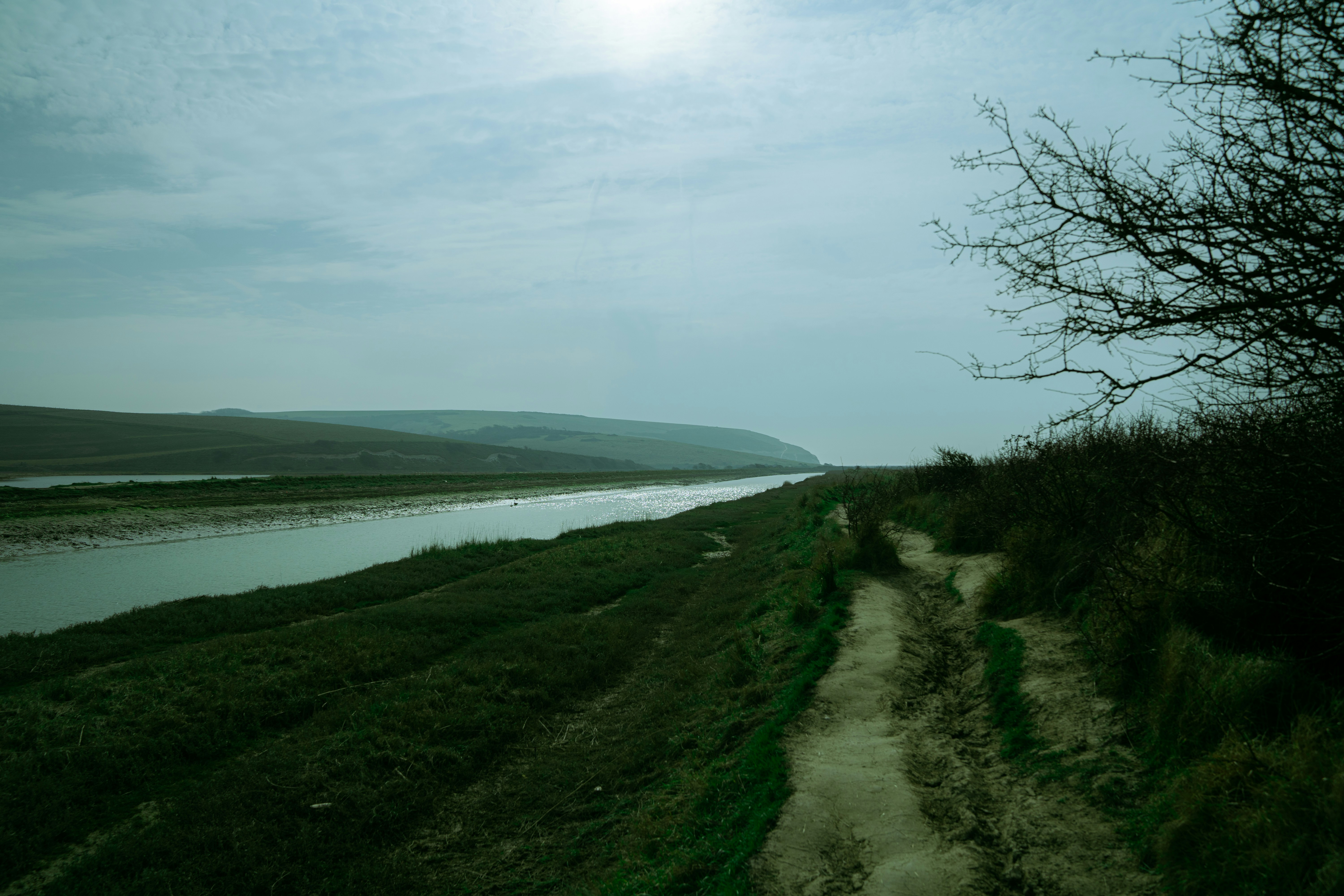 Winding dirt trail beside a reflective river under a cloudy sky.