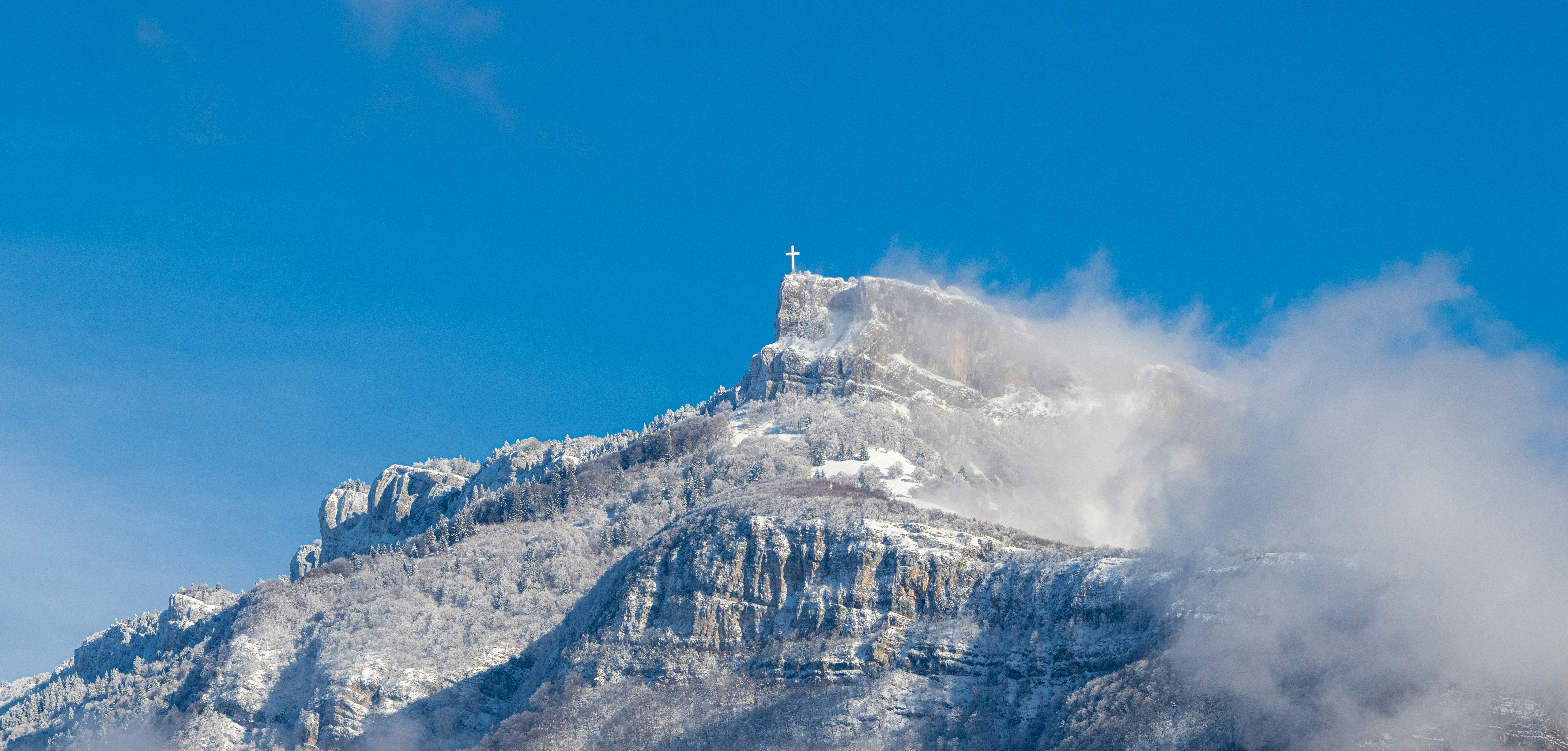 Snow-covered mountain with a cross on the summit beneath a clear blue sky.