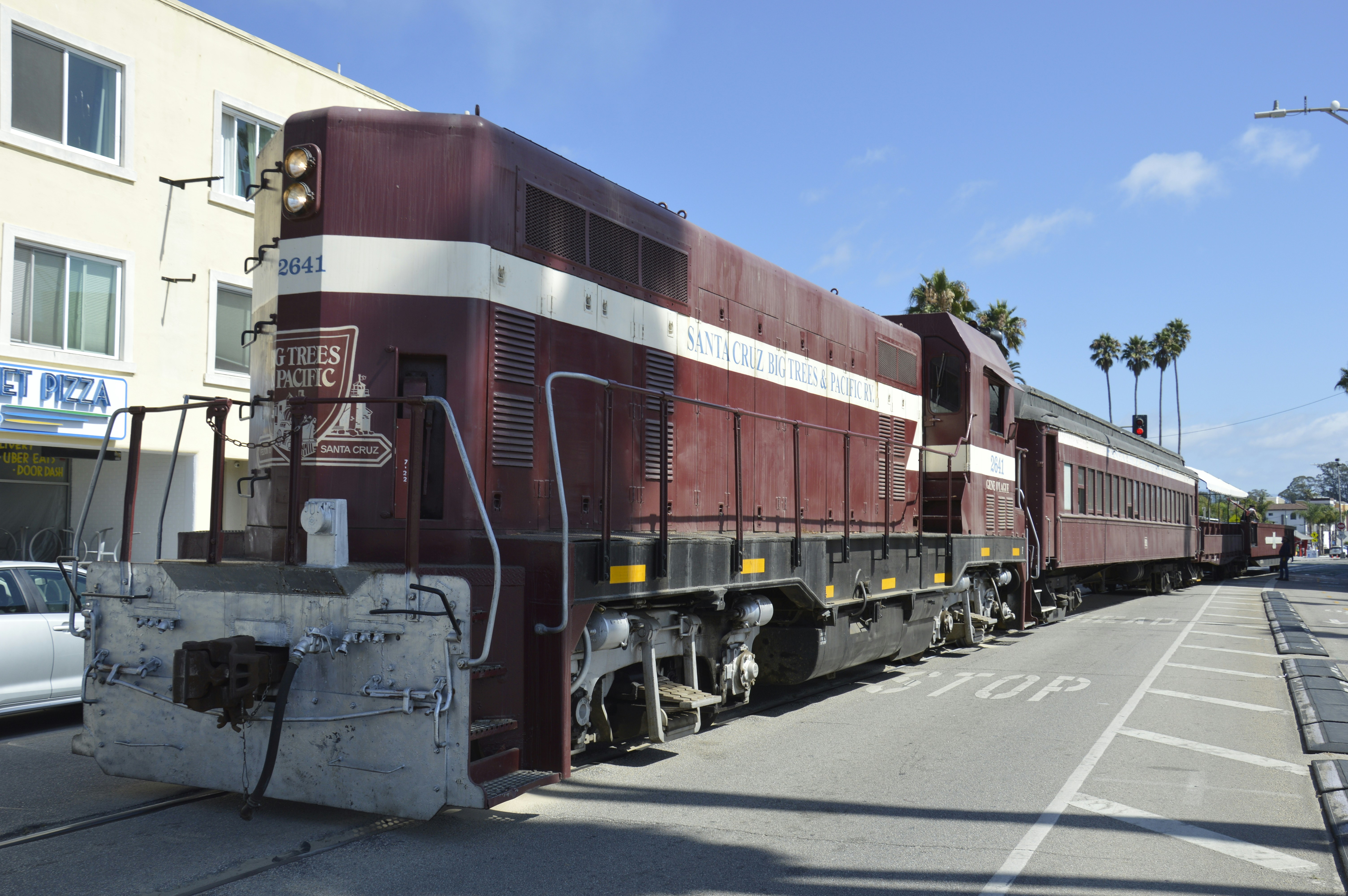 Maroon train stationed on a street with palm trees and buildings under a clear blue sky.