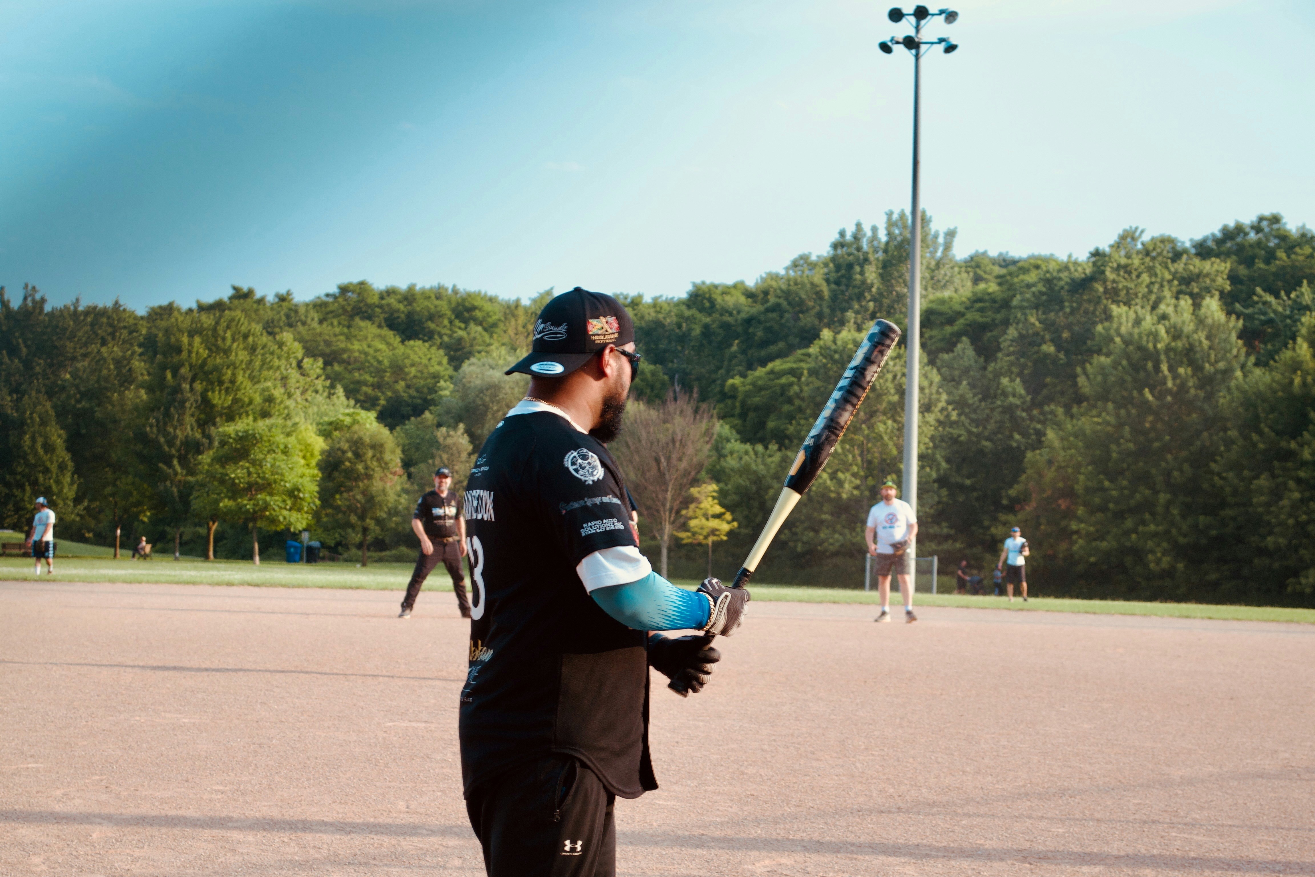 Baseball player holding a bat, poised in a sunlit park with teammates in the background.