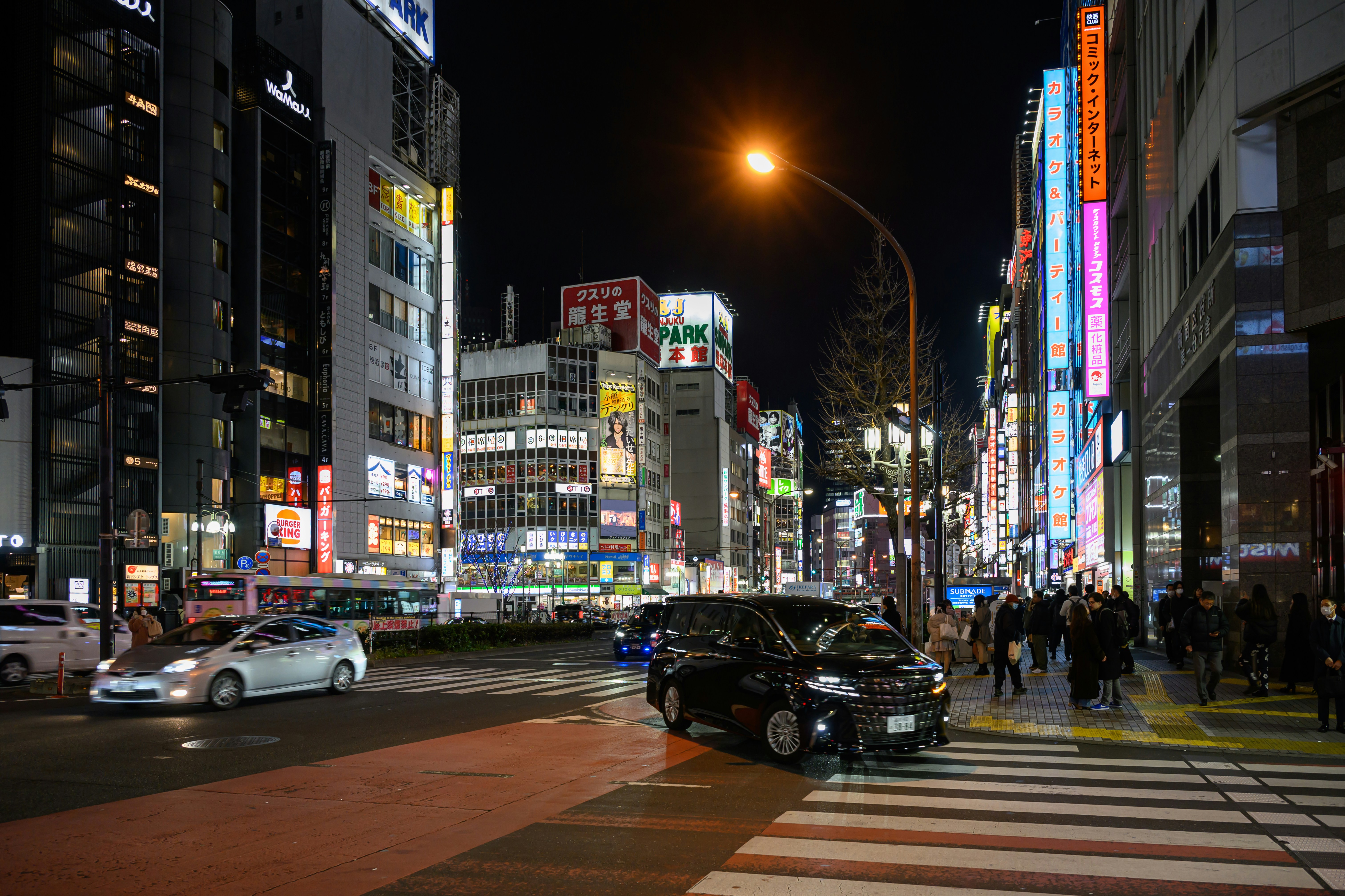 Vibrant city intersection in Shinjuku illuminated by neon signs and bustling with traffic and pedestrians.