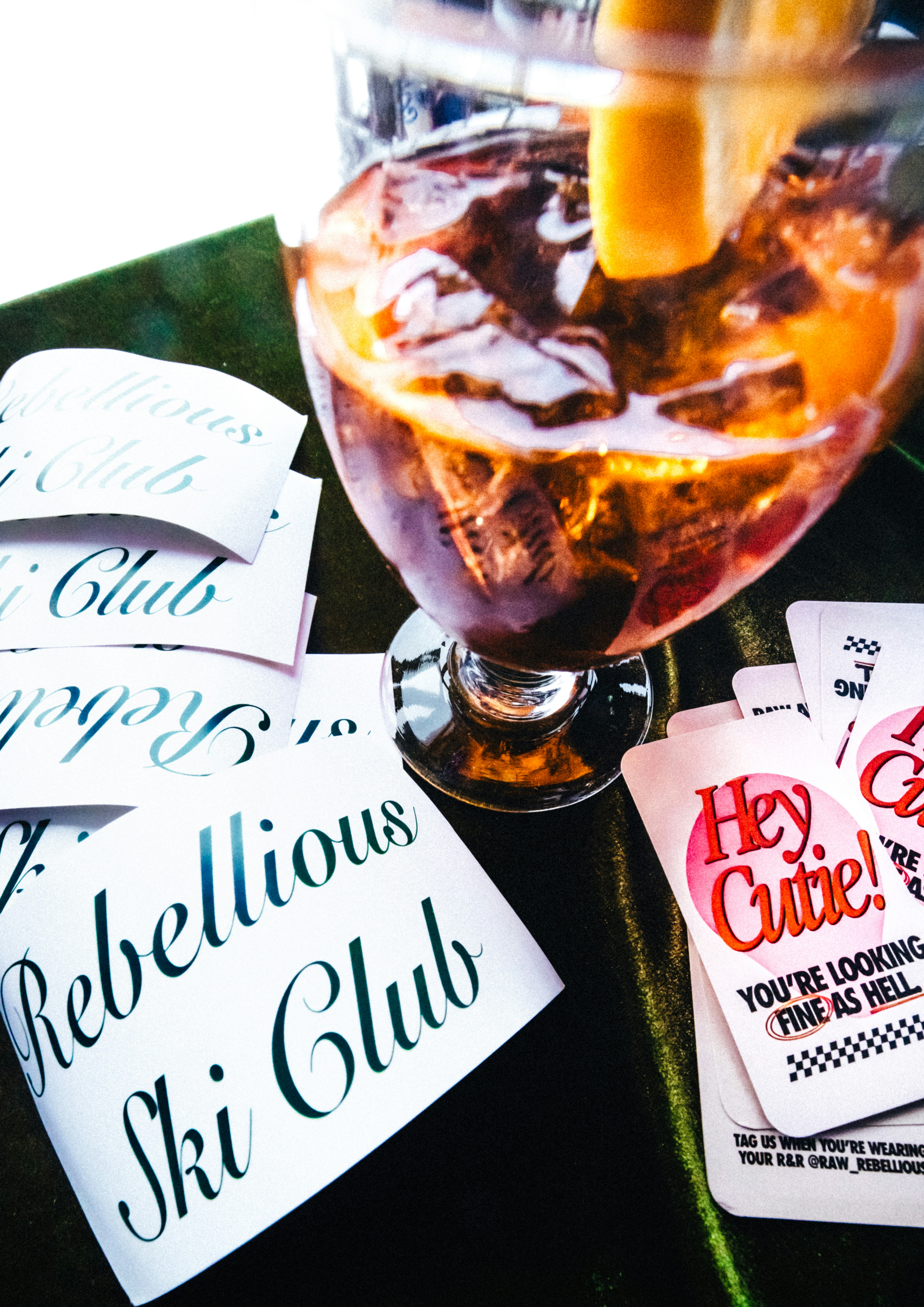 A glass of cocktail garnished with a lemon slice sits atop a green surface, surrounded by playful stickers promoting the 'Rebellious Ski Club' and other fun messages.