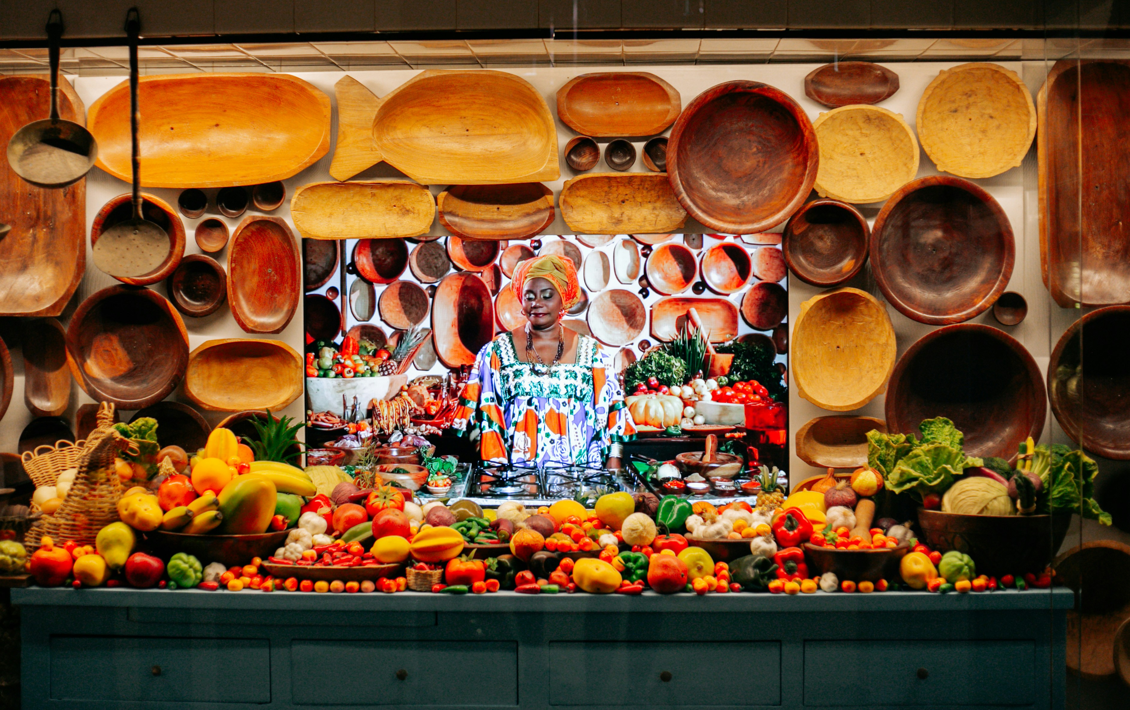Woman in colorful attire stands before an array of fruits and woven baskets.