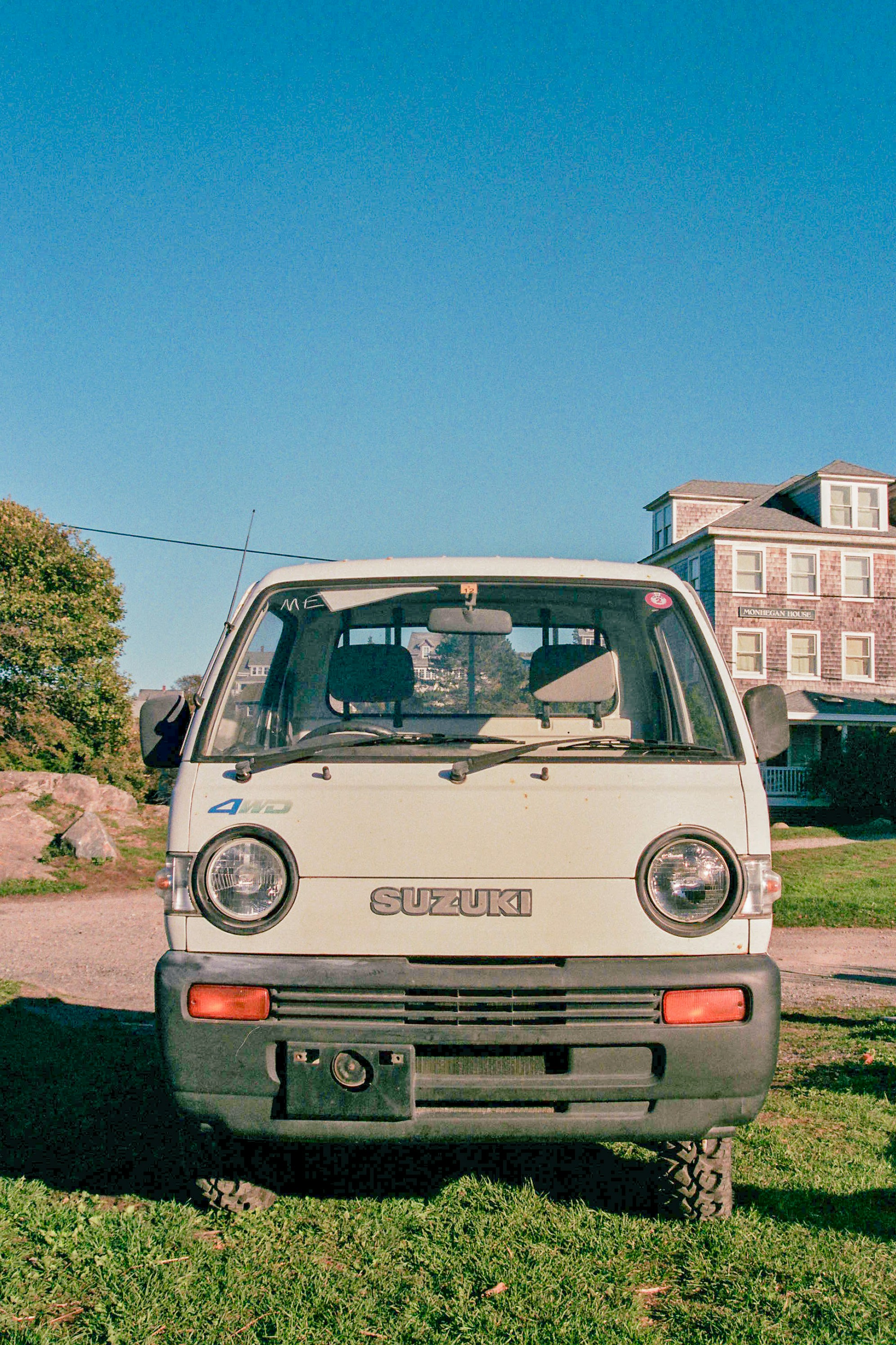 Front-facing white Suzuki van parked on a grassy lot beneath a clear blue sky, with a suburban house in the background.