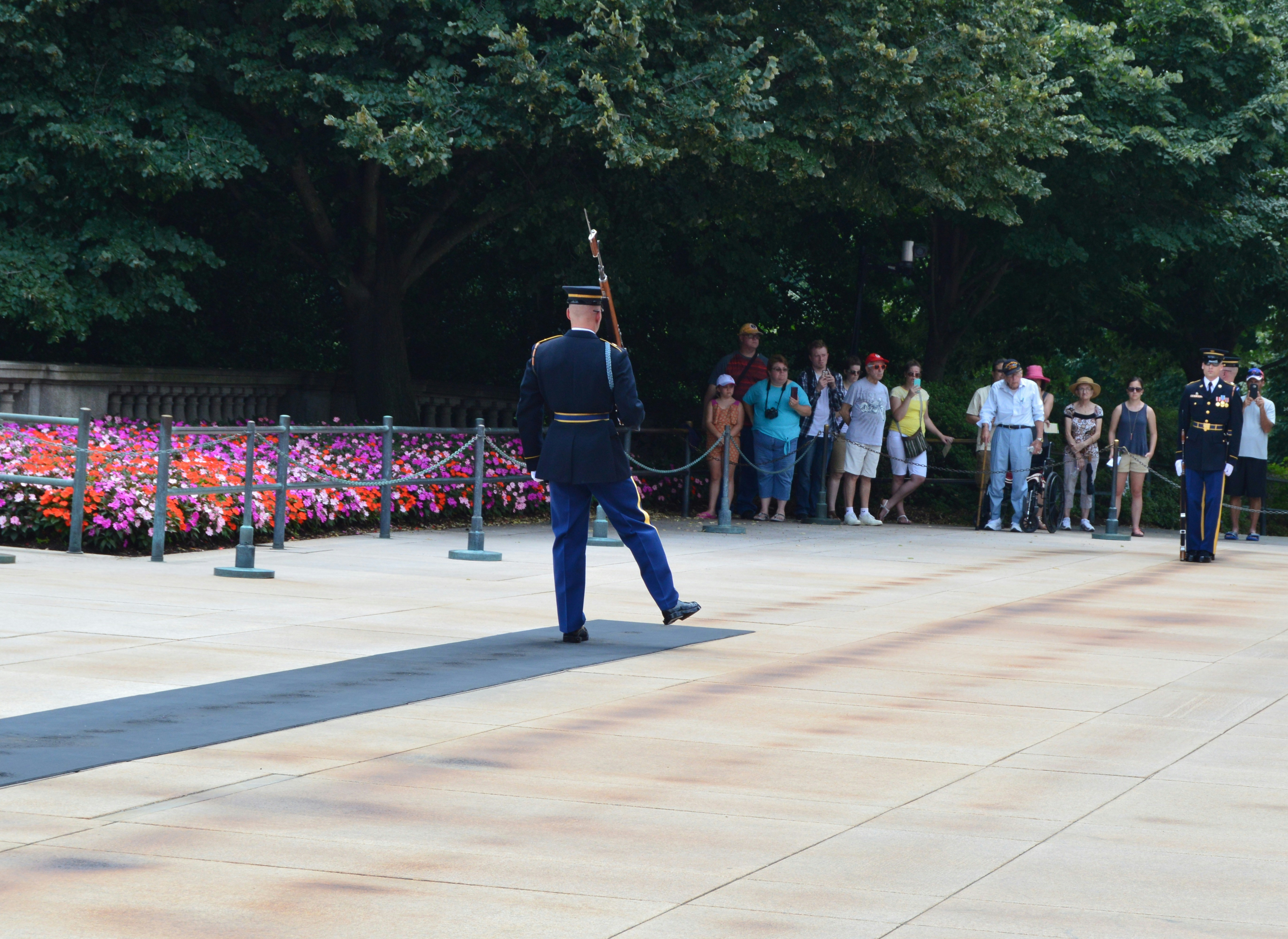 Soldier in uniform marches with a rifle as a crowd observes at a memorial site.