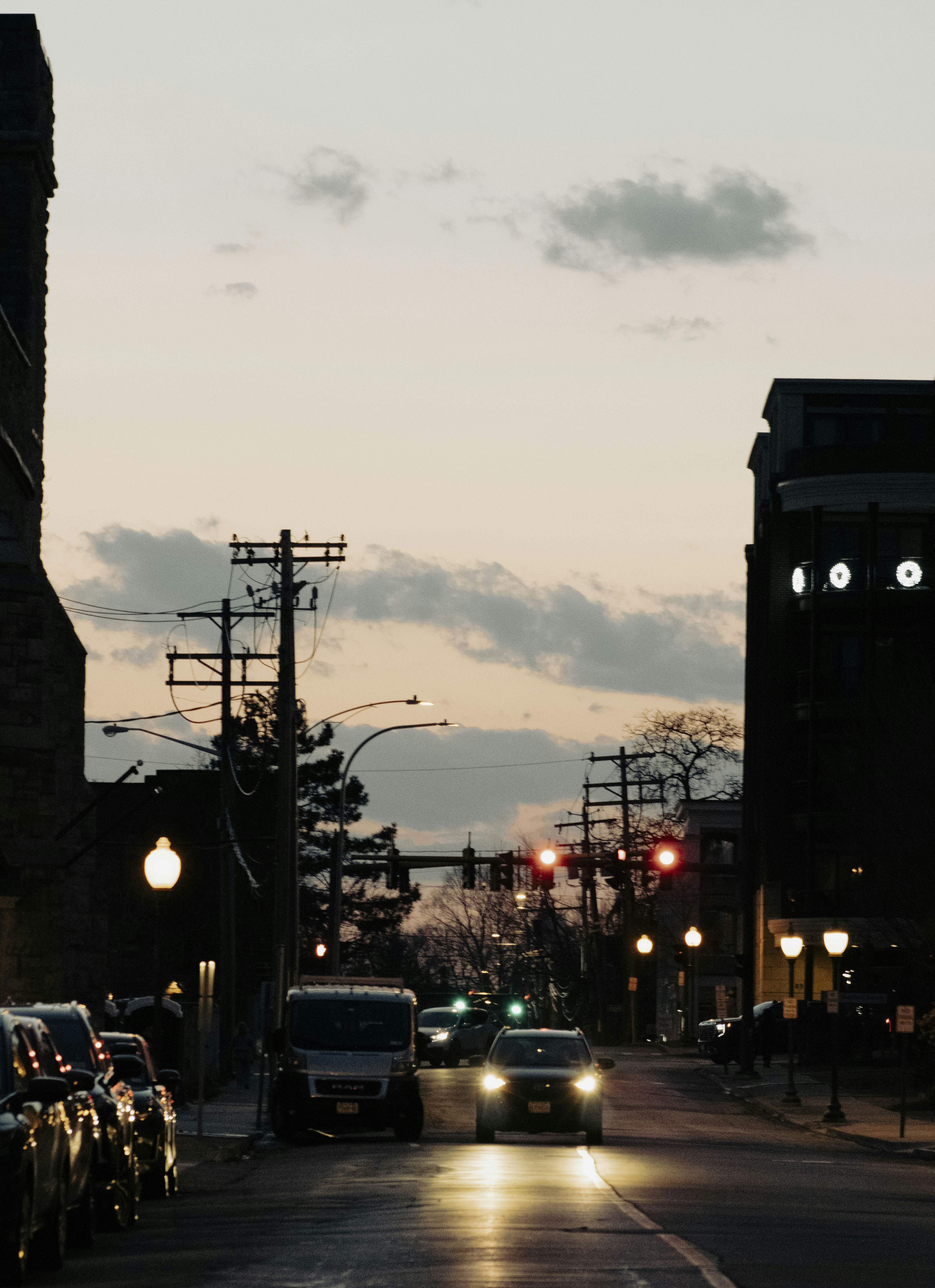Dimly lit street scene at twilight, featuring a car approaching traffic lights amidst silhouetted buildings and power lines.