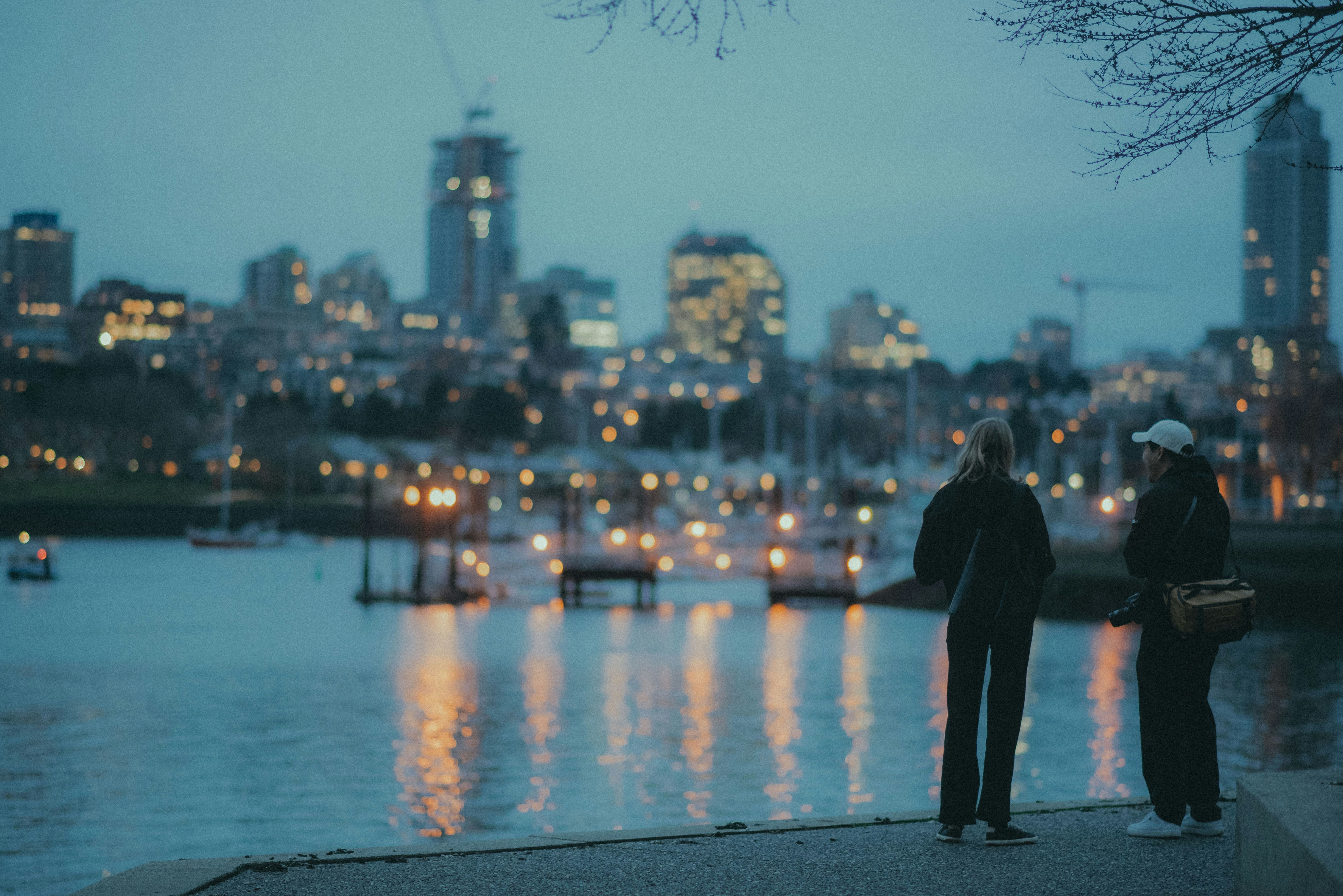 Two people converse near a waterfront with city lights reflecting on the water at dusk.