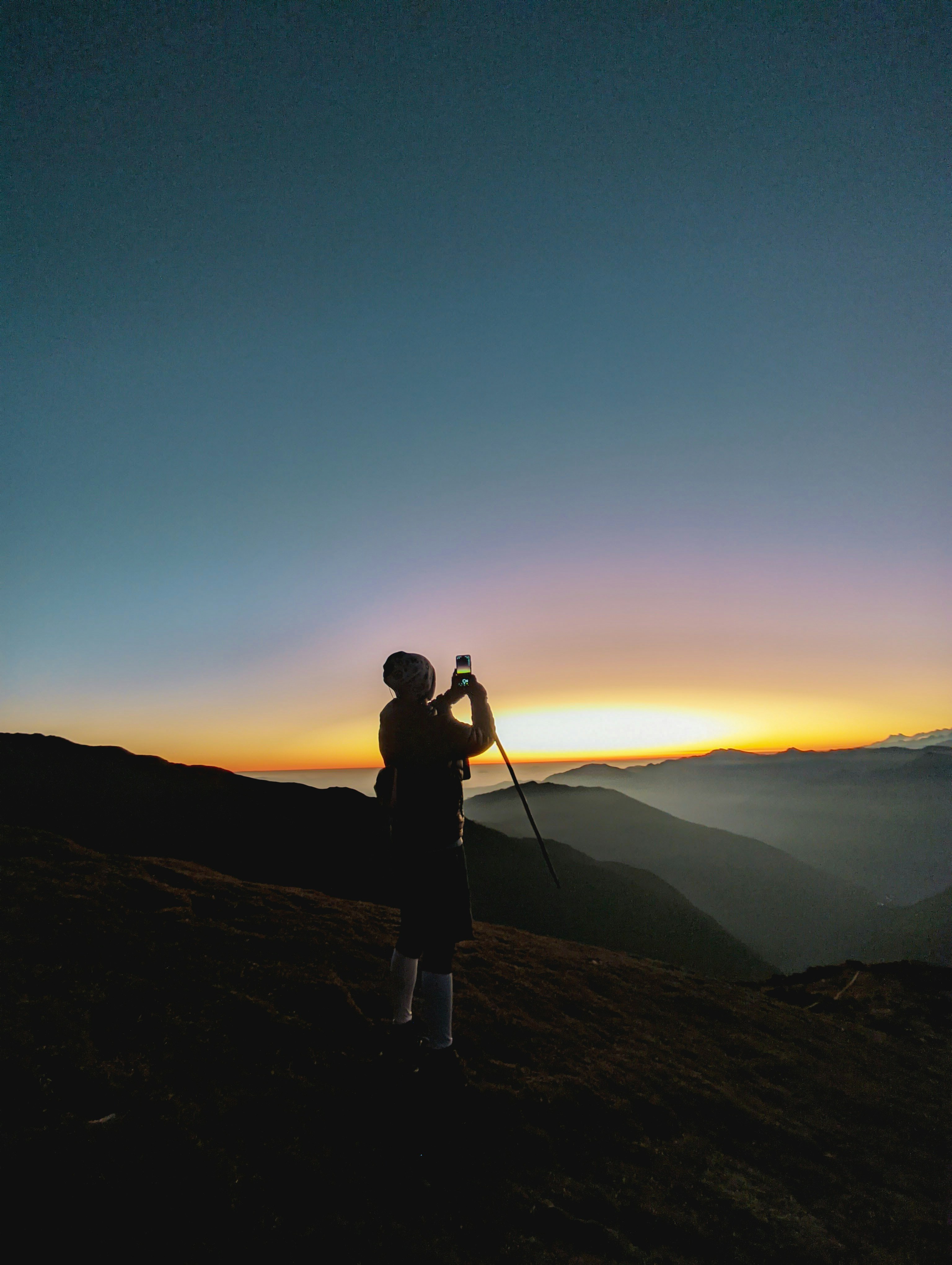 Silhouette of a photographer on a ridge at sunset, camera on a tripod angled toward the glowing horizon over layered mountains.